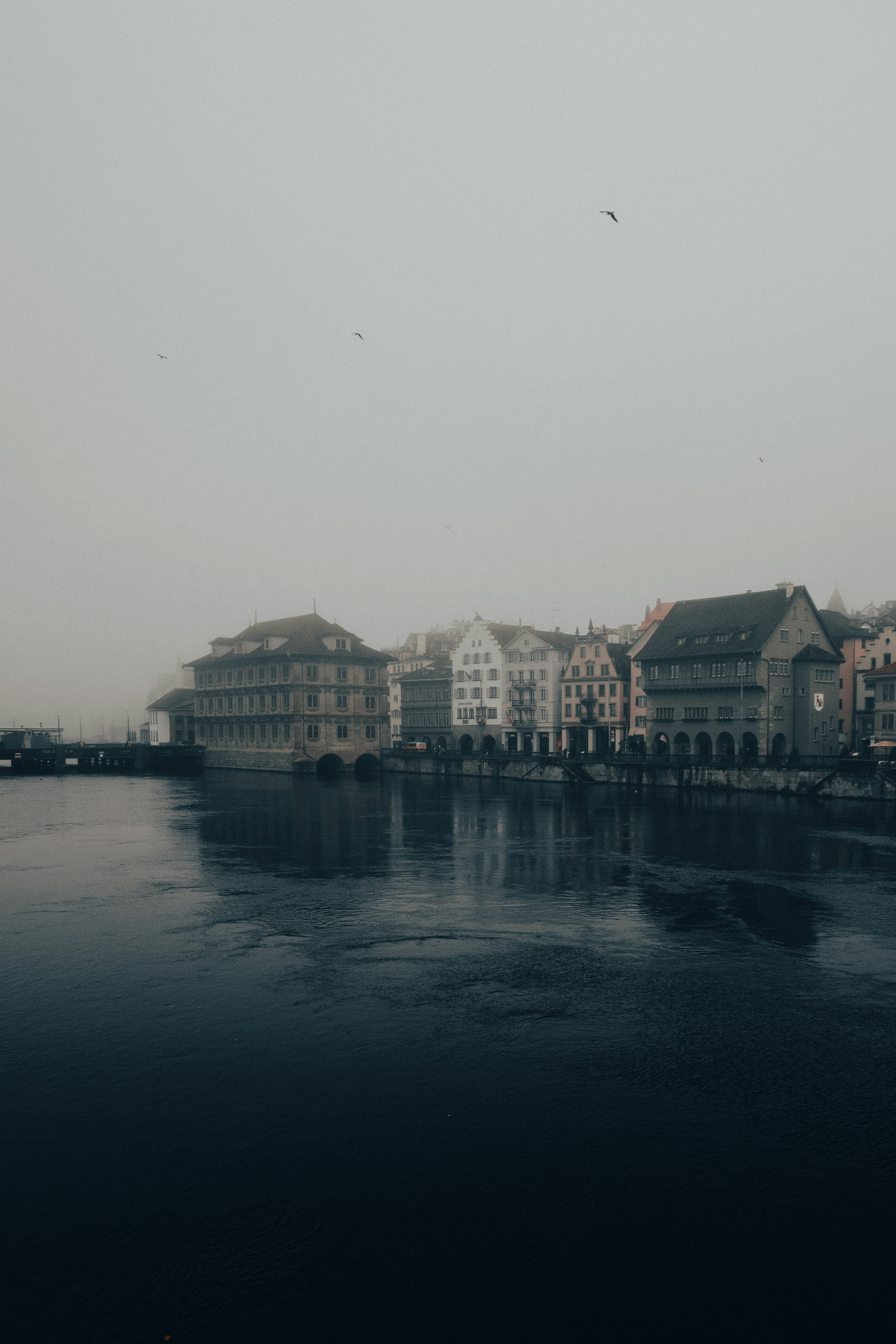 Foggy riverside scene with historic buildings reflected in calm water.