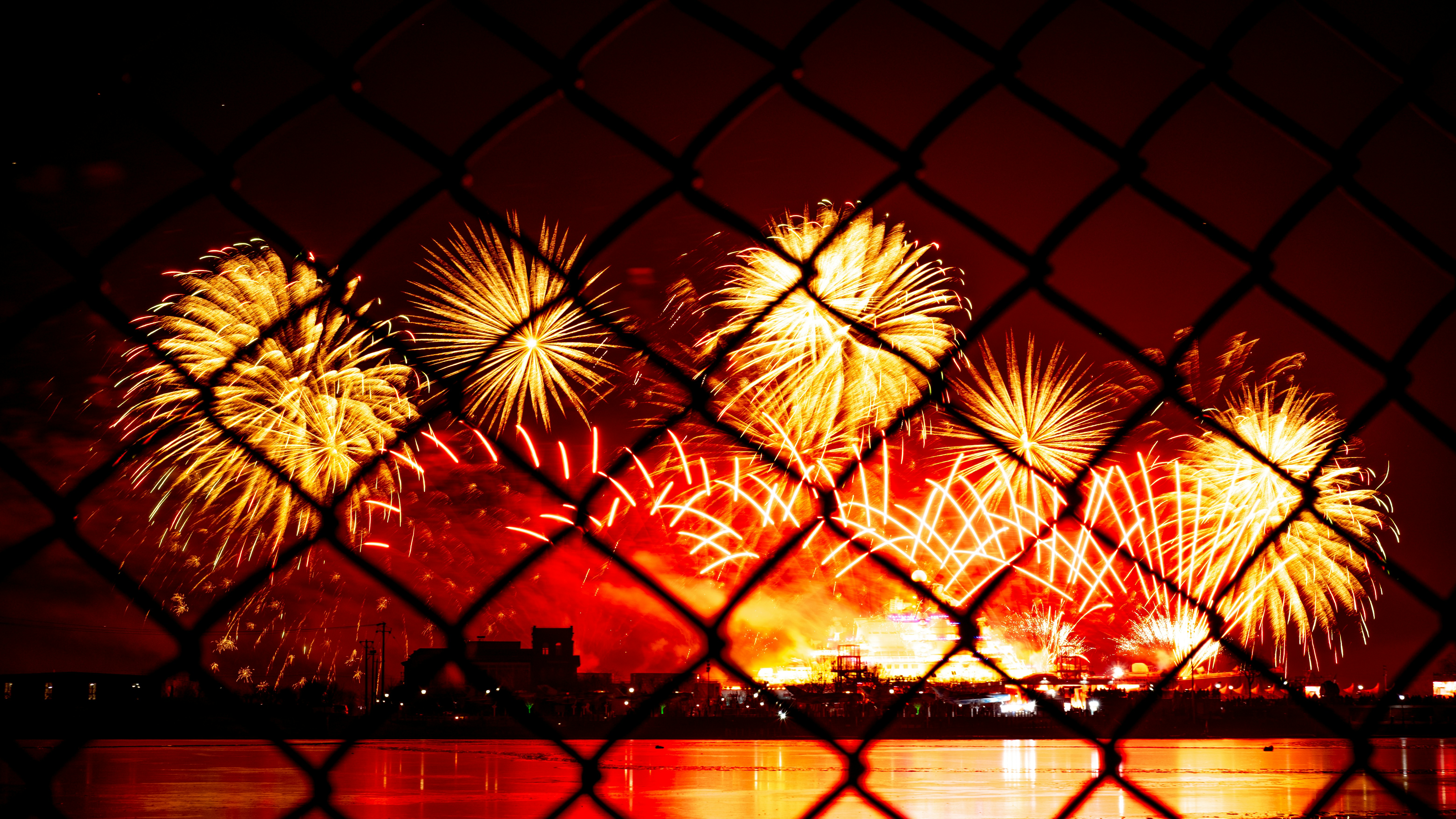 Framed Nightburst: Fireworks Over a Waterfront City Through a Chain-Link Veil