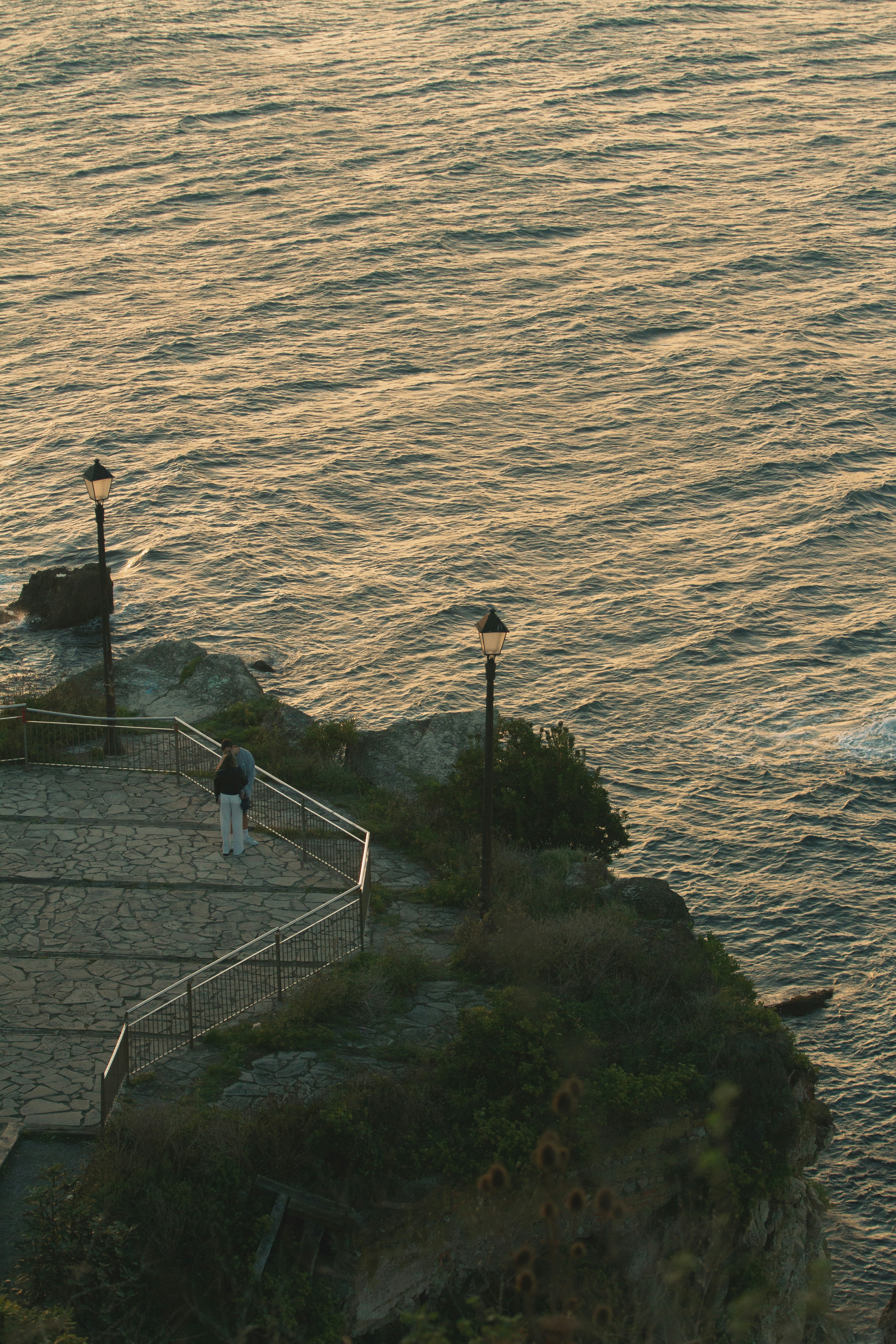 Person standing on a stone terrace overlooking a calm sea at sunset.
