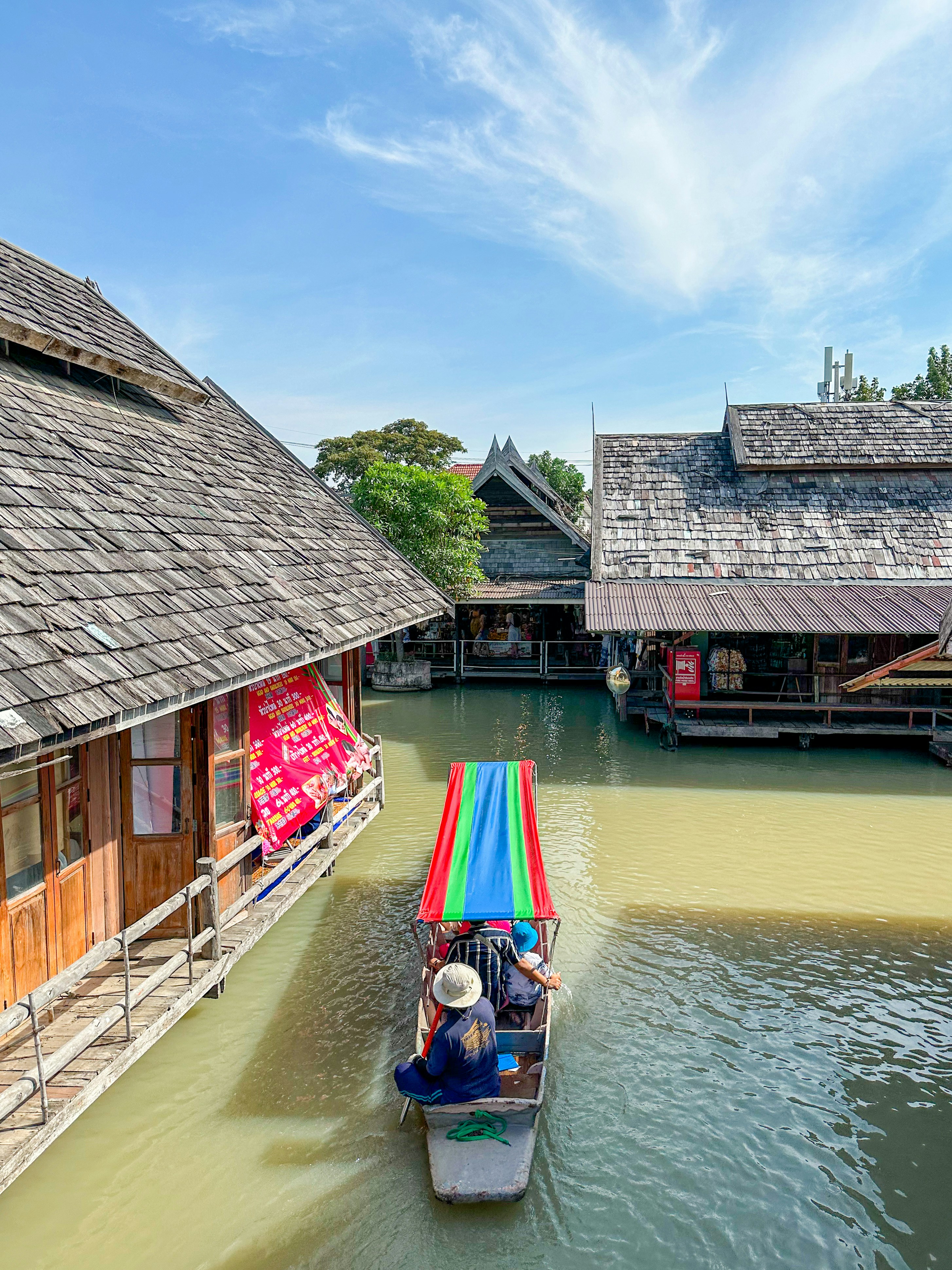 Boat with colorful canopy glides through narrow canal between wooden market buildings under a clear sky.