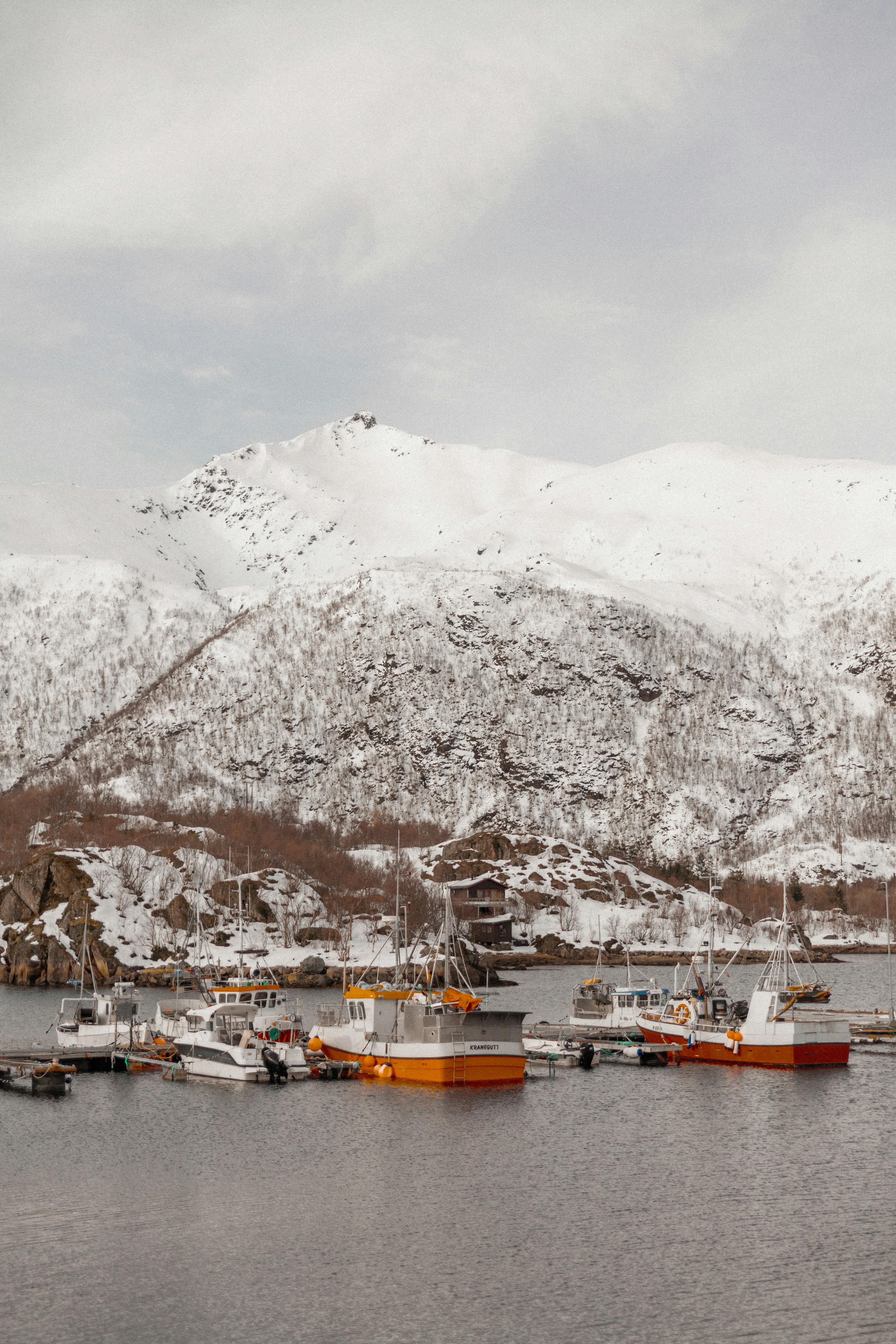 Boats docked in a calm harbor beneath snow-covered mountains on a cloudy day.