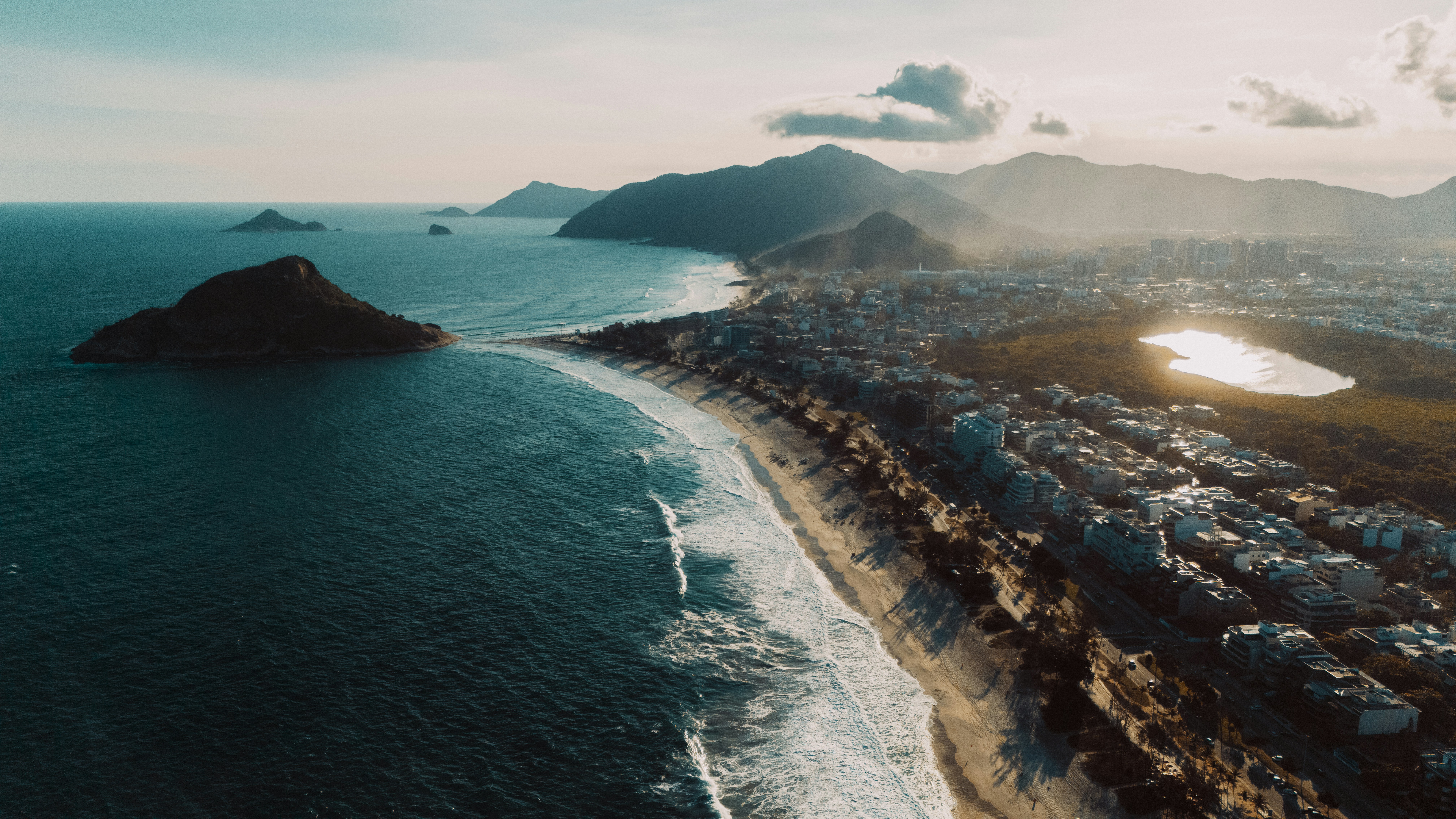 Aerial view of Recreio Beach at sunset, with waves crashing along the shore and mountains in the background.