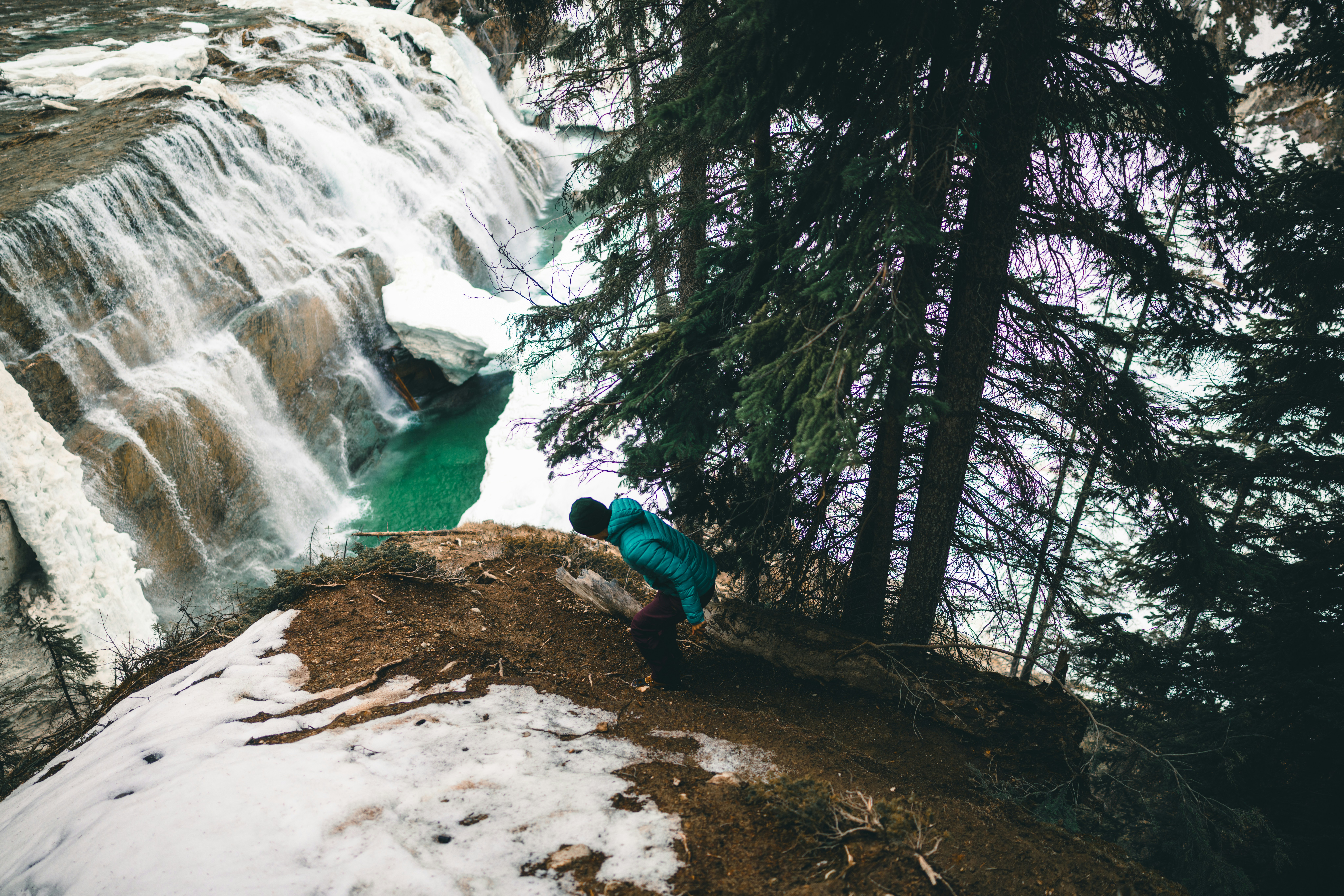 Person in a teal jacket stands near a snowy edge overlooking a cascading waterfall surrounded by evergreens.