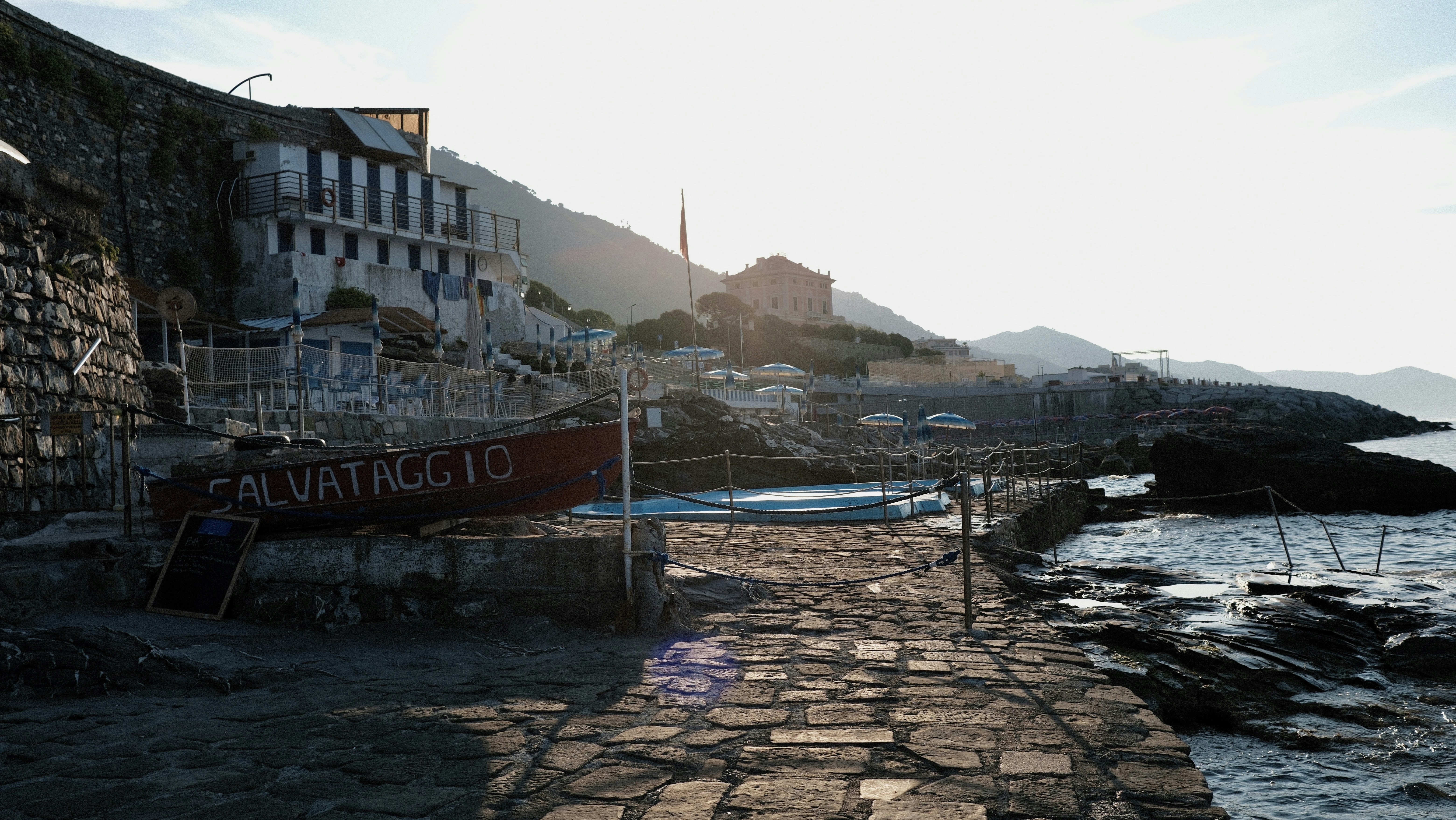 Stone walkway along a tranquil coastline at sunset, with mountains and rustic buildings in the background.