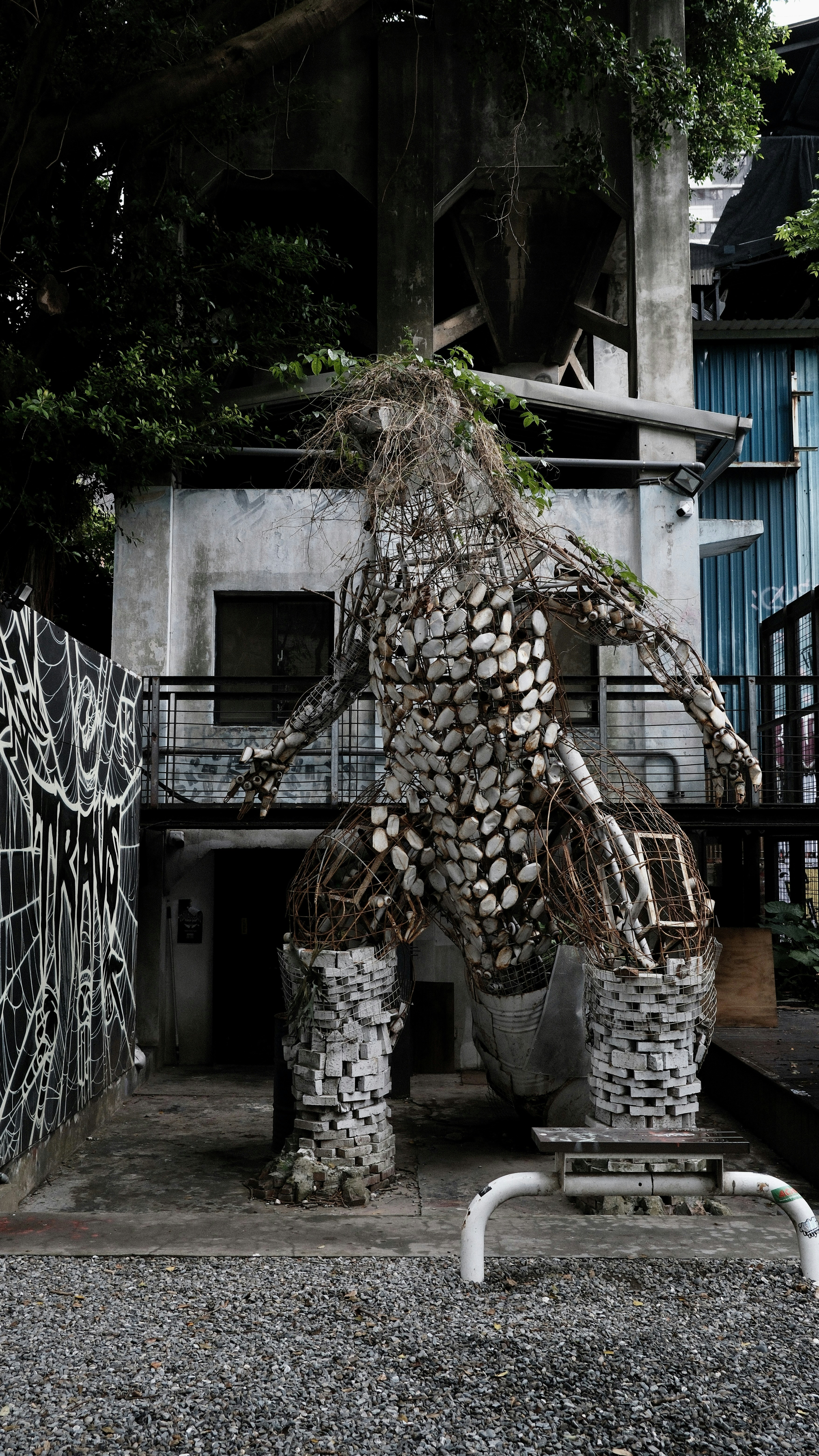 Sculpture of a humanoid figure made from logs and branches stands in front of an industrial-style building.