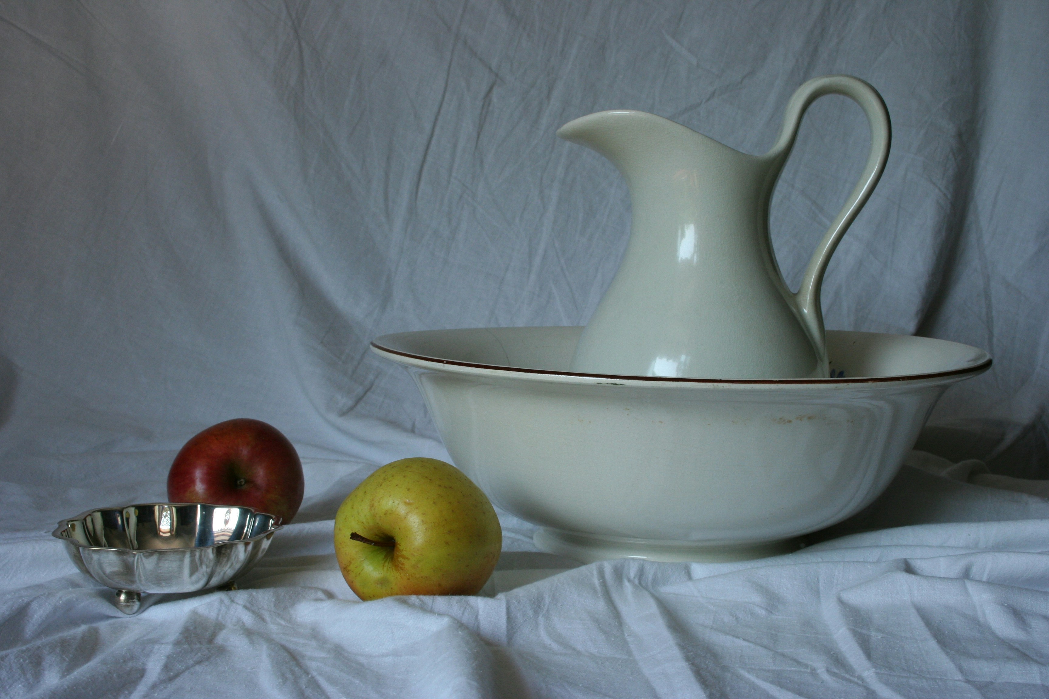 Porcelain pitcher and bowl with red and yellow apples on a draped fabric backdrop.