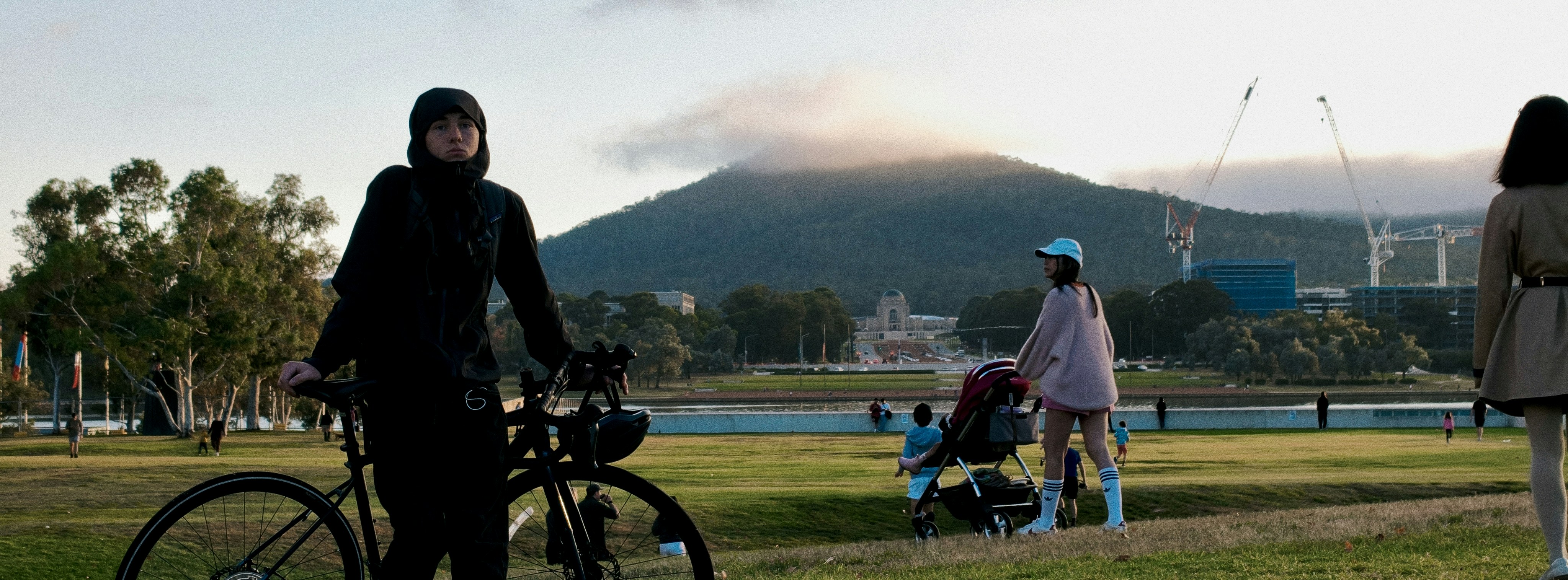 People strolling and cycling near a lake with Mount Ainslie and city skyline in the background.