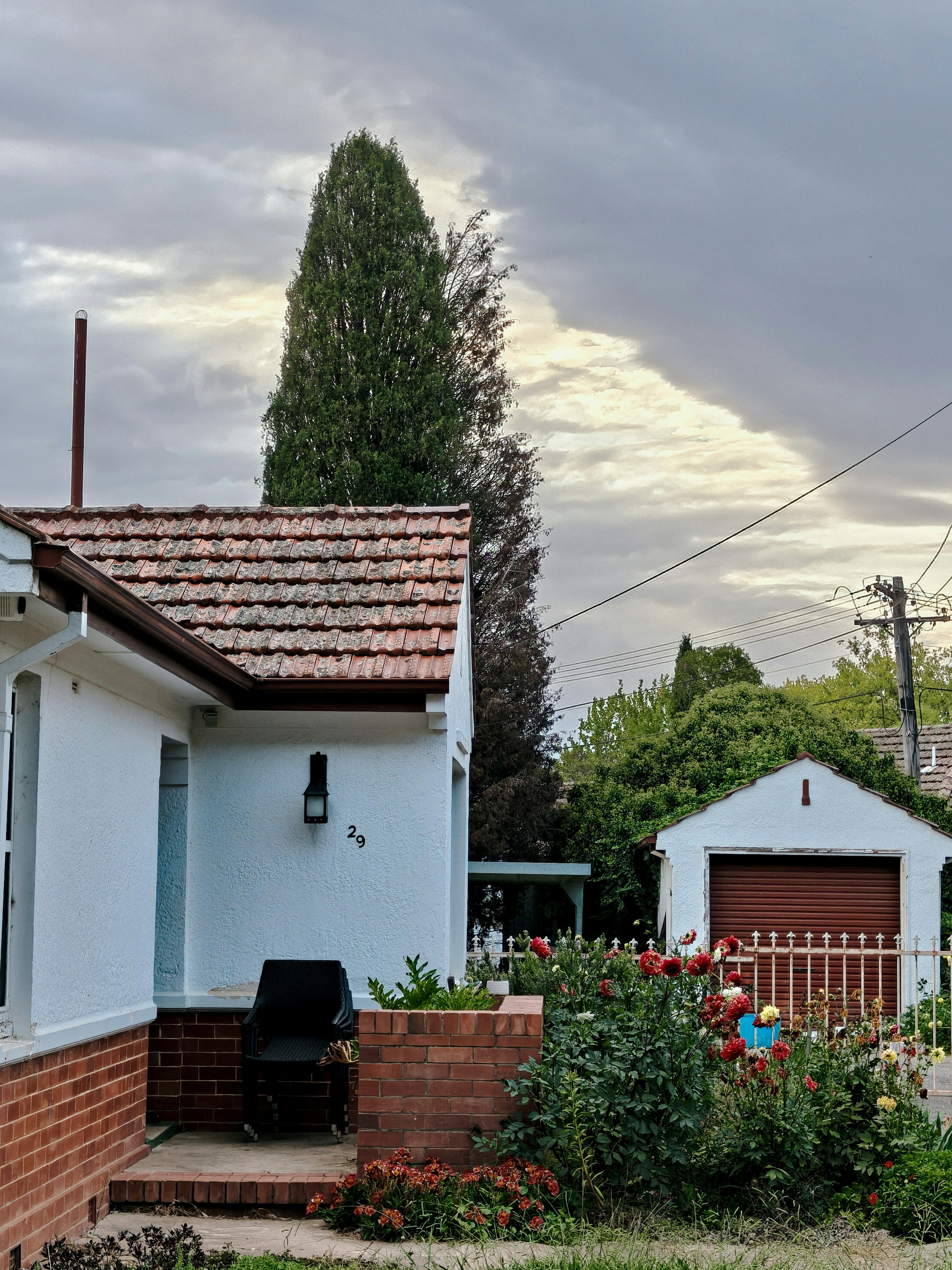 A house with a garden featuring colorful flowers and a tall tree, set against a cloudy sky, creating a calm suburban atmosphere.