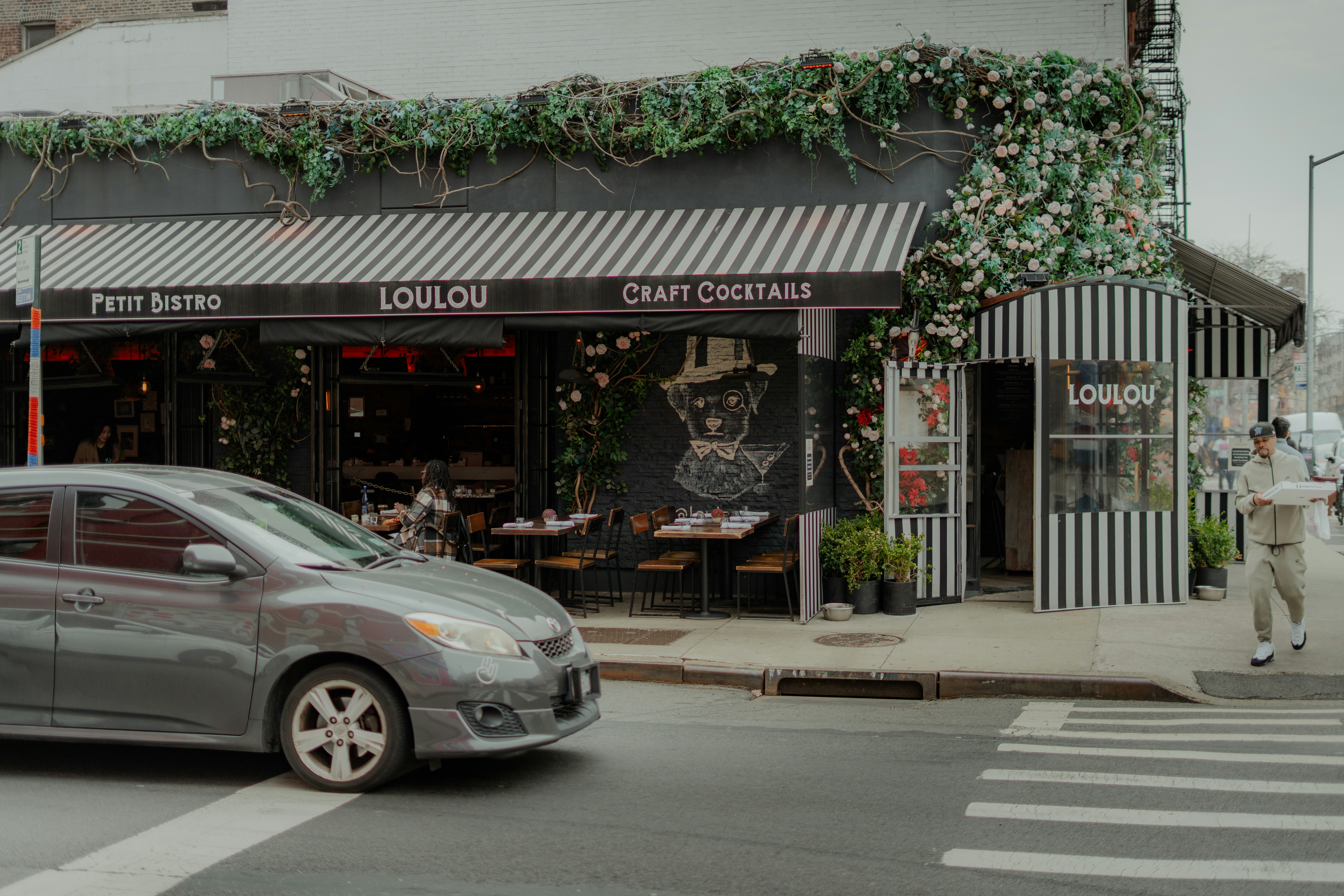 Street view of a car passing by a cafe with striped awnings and lush greenery on the facade.