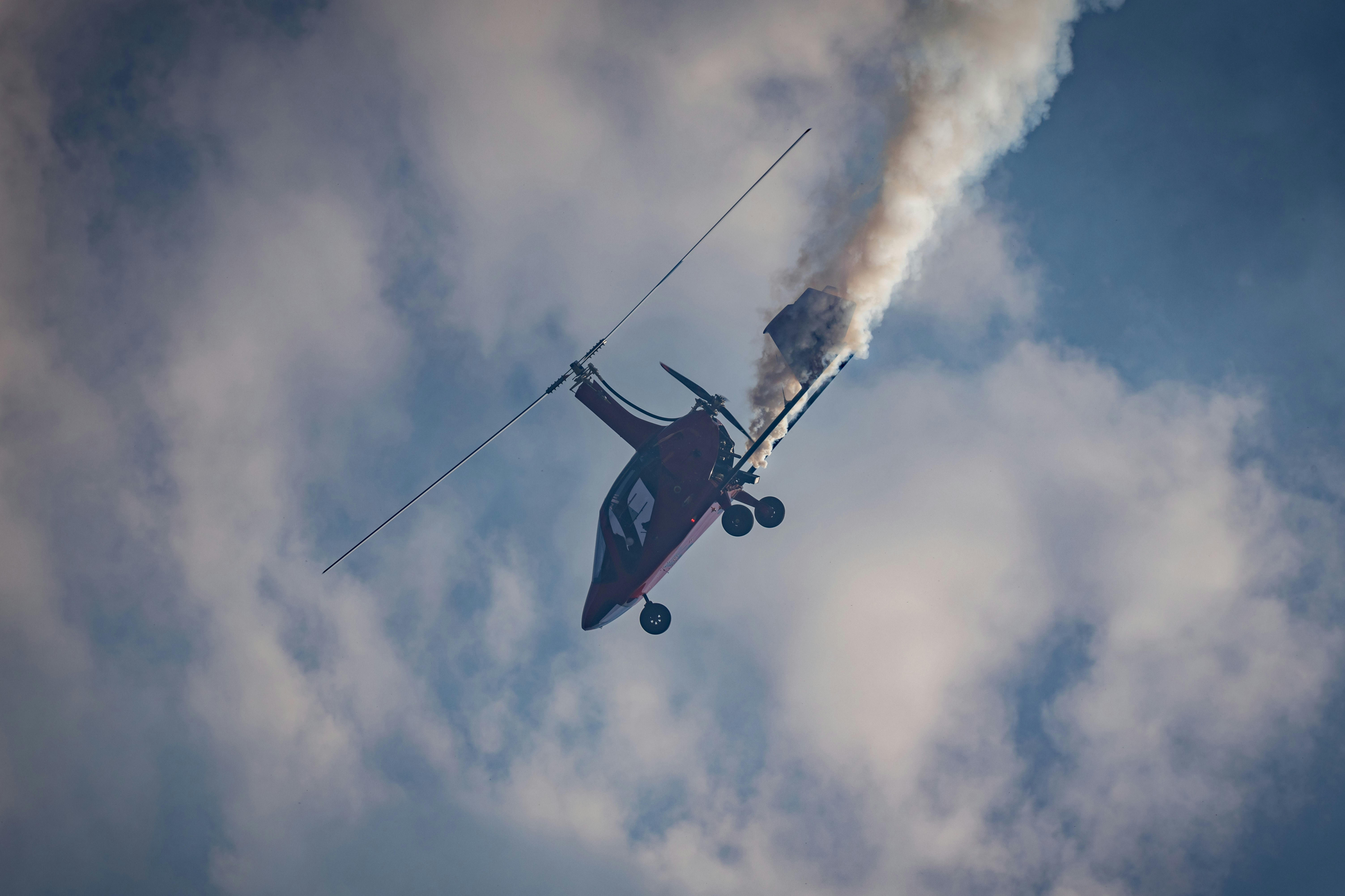 Red aerobatic aircraft threads a plume of smoke through a cloud-dappled sky