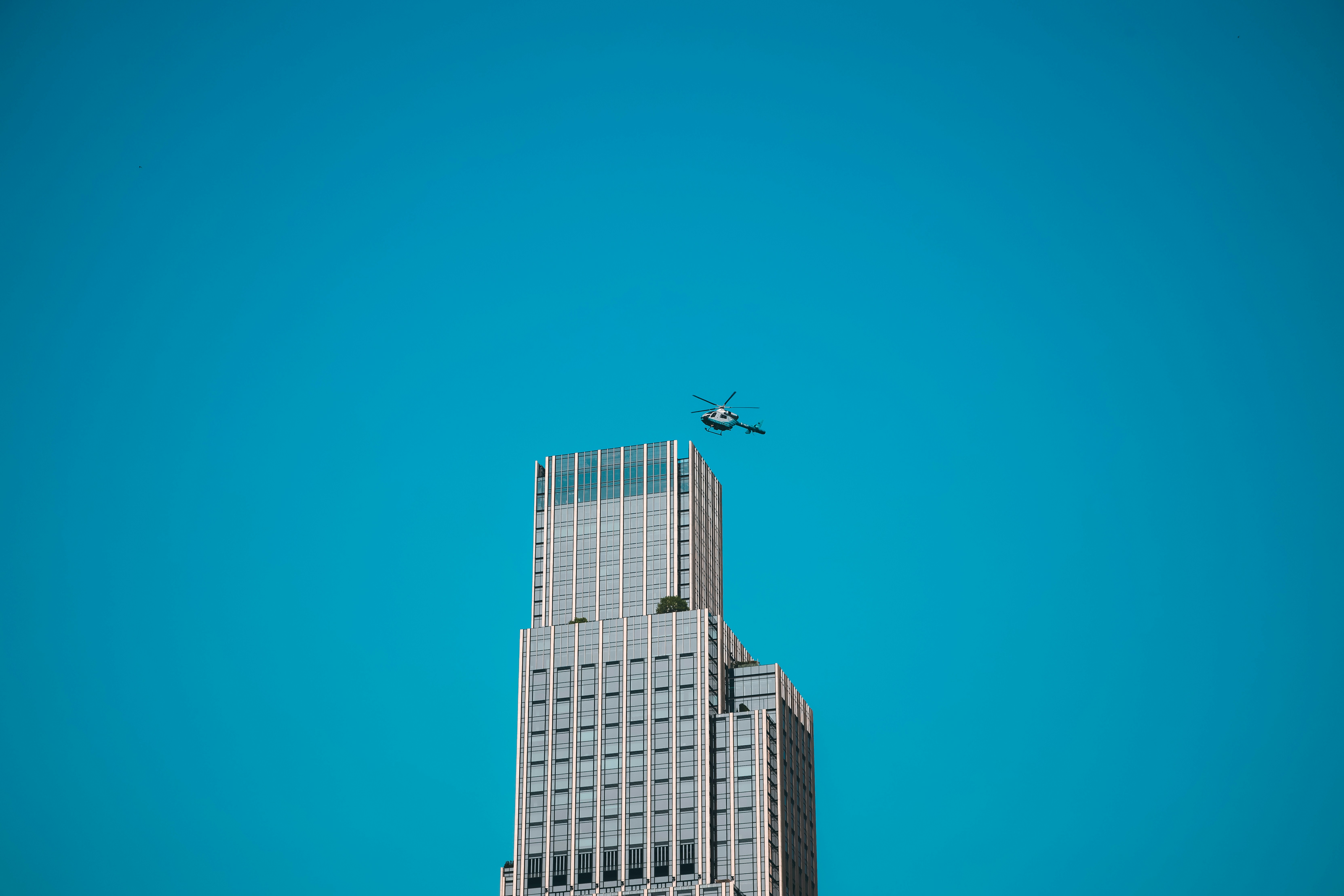 Helicopter flying above a modern skyscraper against a clear blue sky.