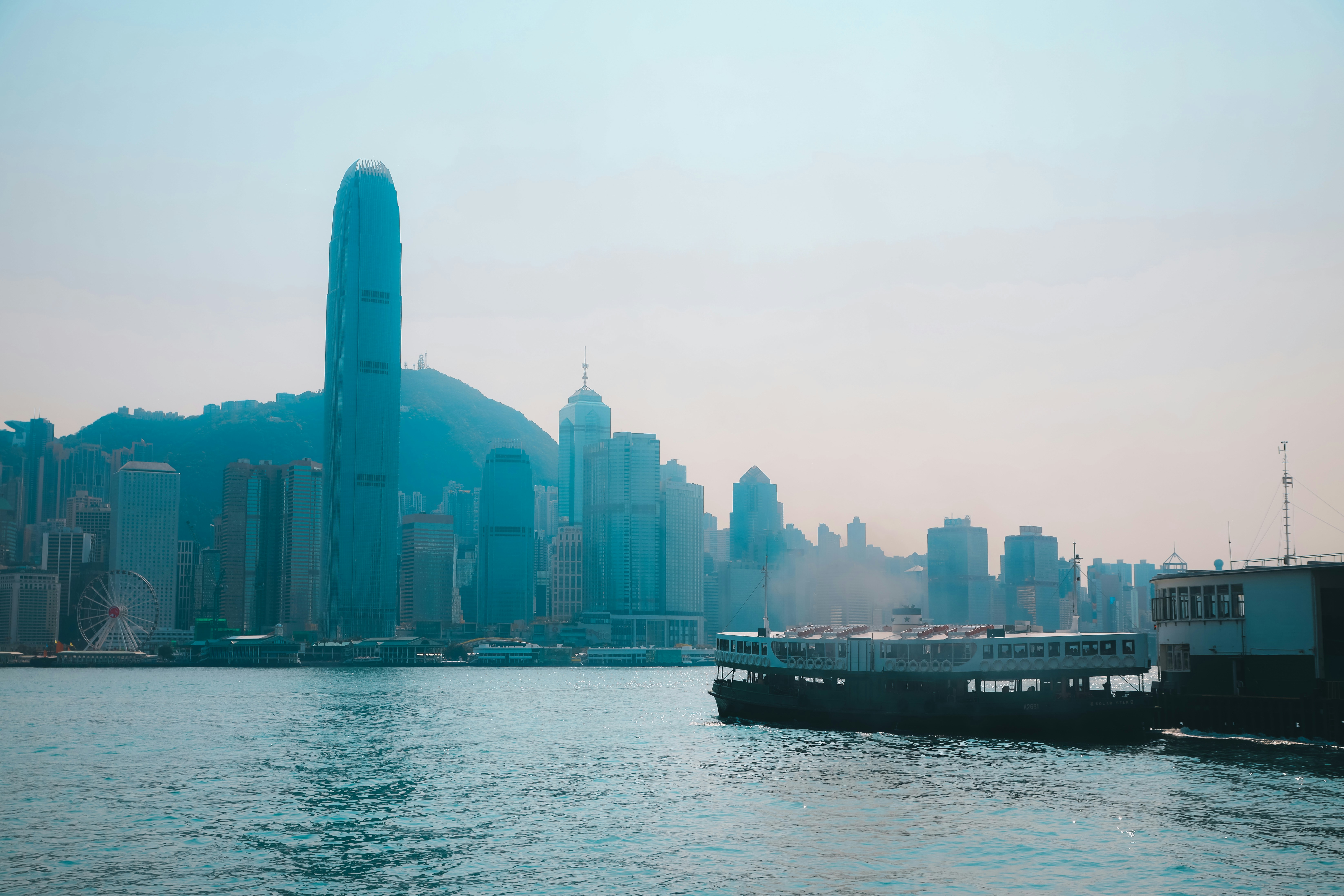 Ferry navigating the waters with the Hong Kong skyline towering in the background under a hazy sky.