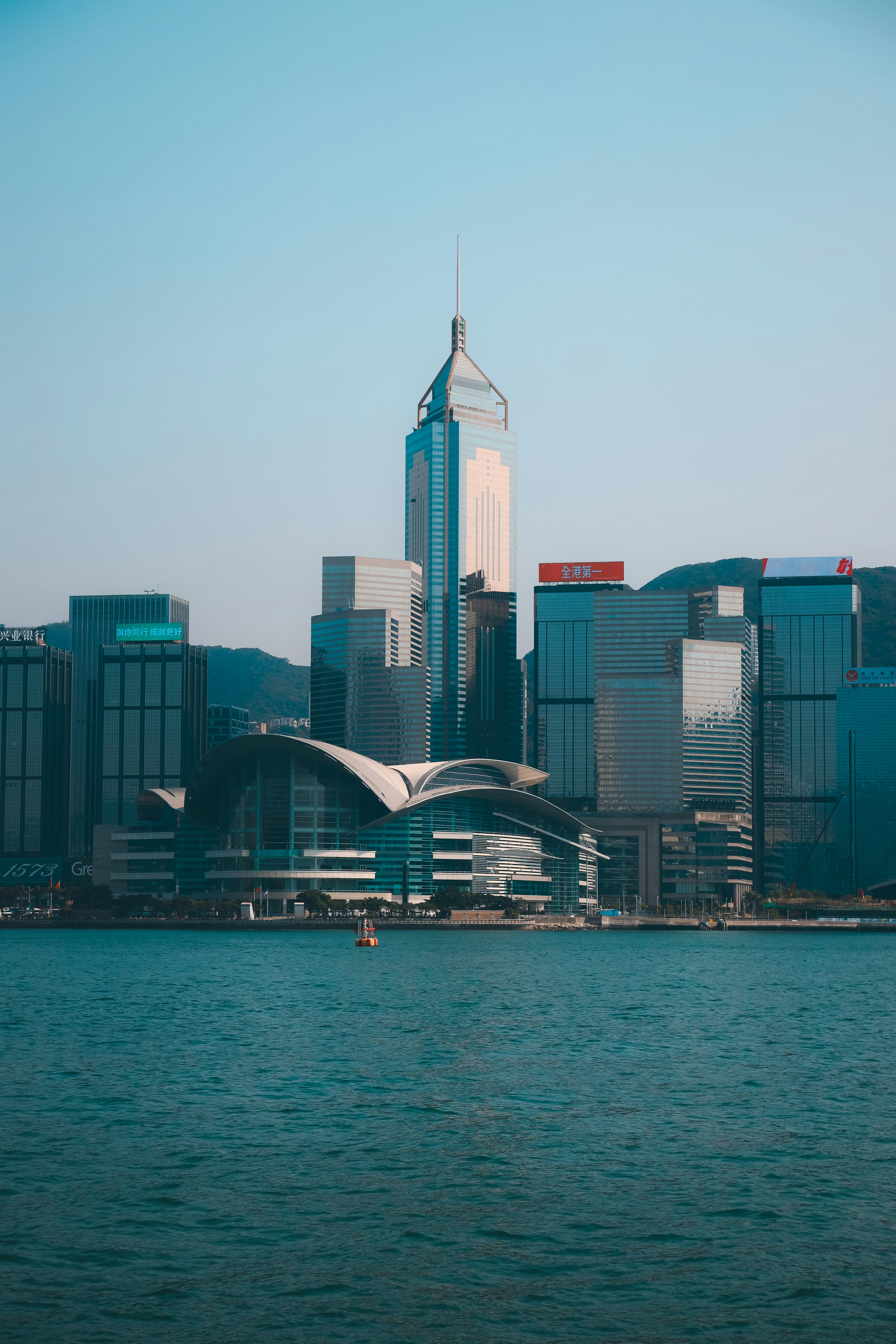 Hong Kong skyscrapers rise against a clear blue sky, with a tranquil harbor in the foreground.