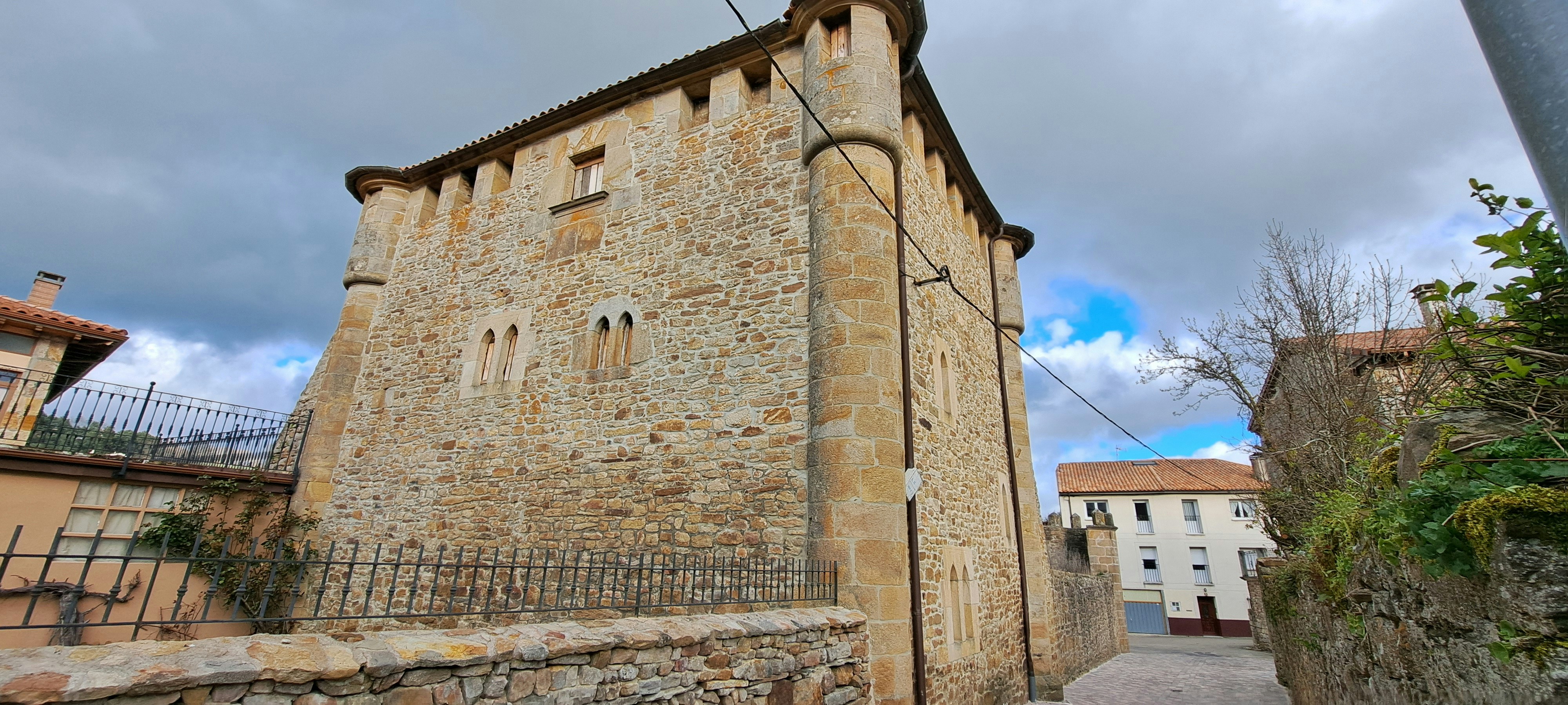 Stone tower with cylindrical turrets set against a partly cloudy sky and adjacent modern buildings.