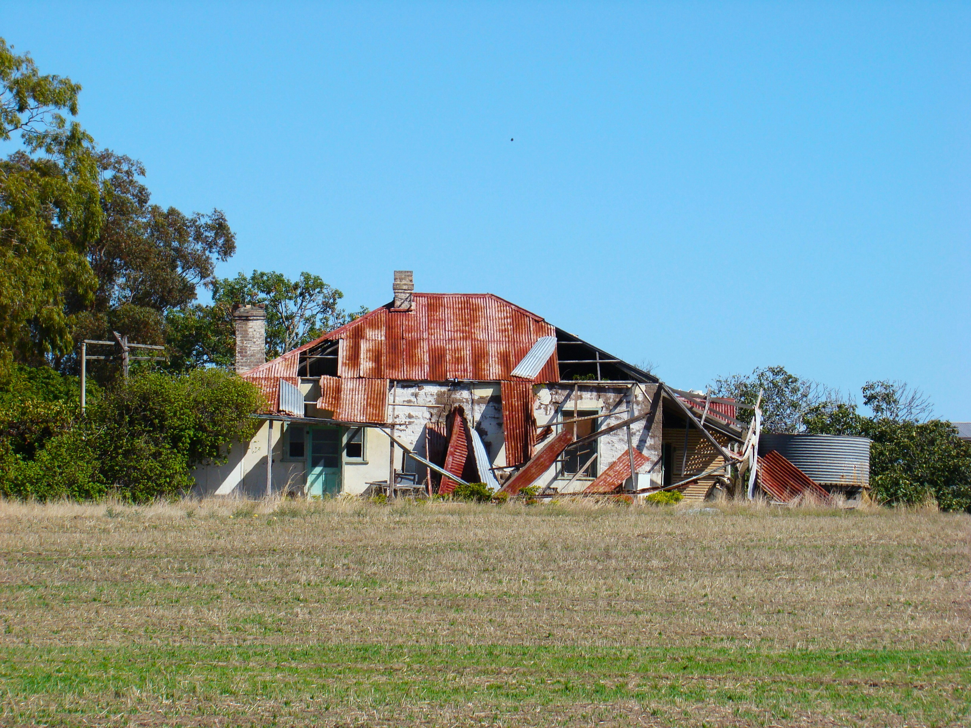 Weathered barn remnant with rusted corrugated metal and collapsed sections sits in a grassy field beneath a clear blue sky. Rural landscape photograph.