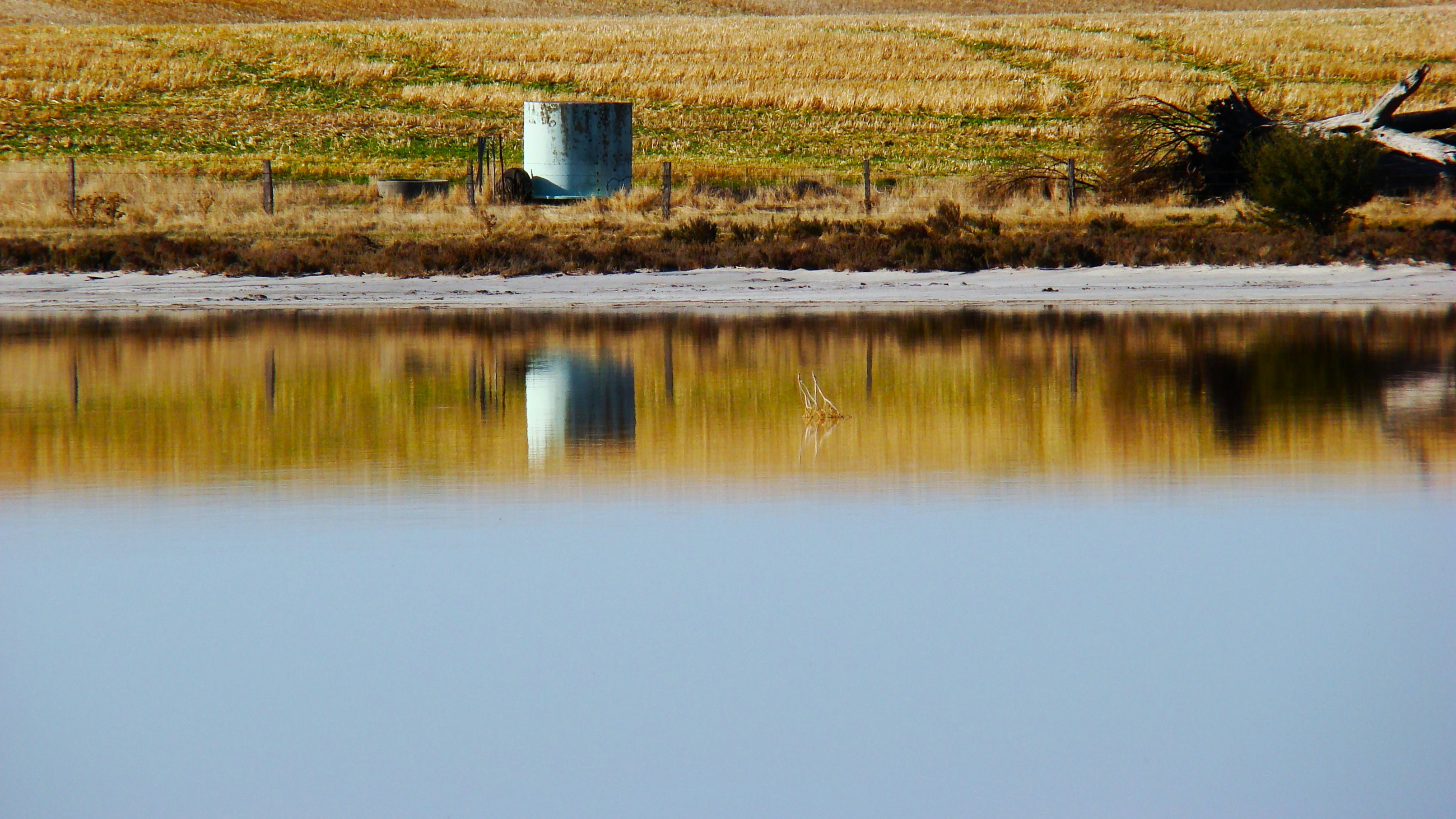 Metal water tank reflecting in a calm lake surrounded by grassy landscape.