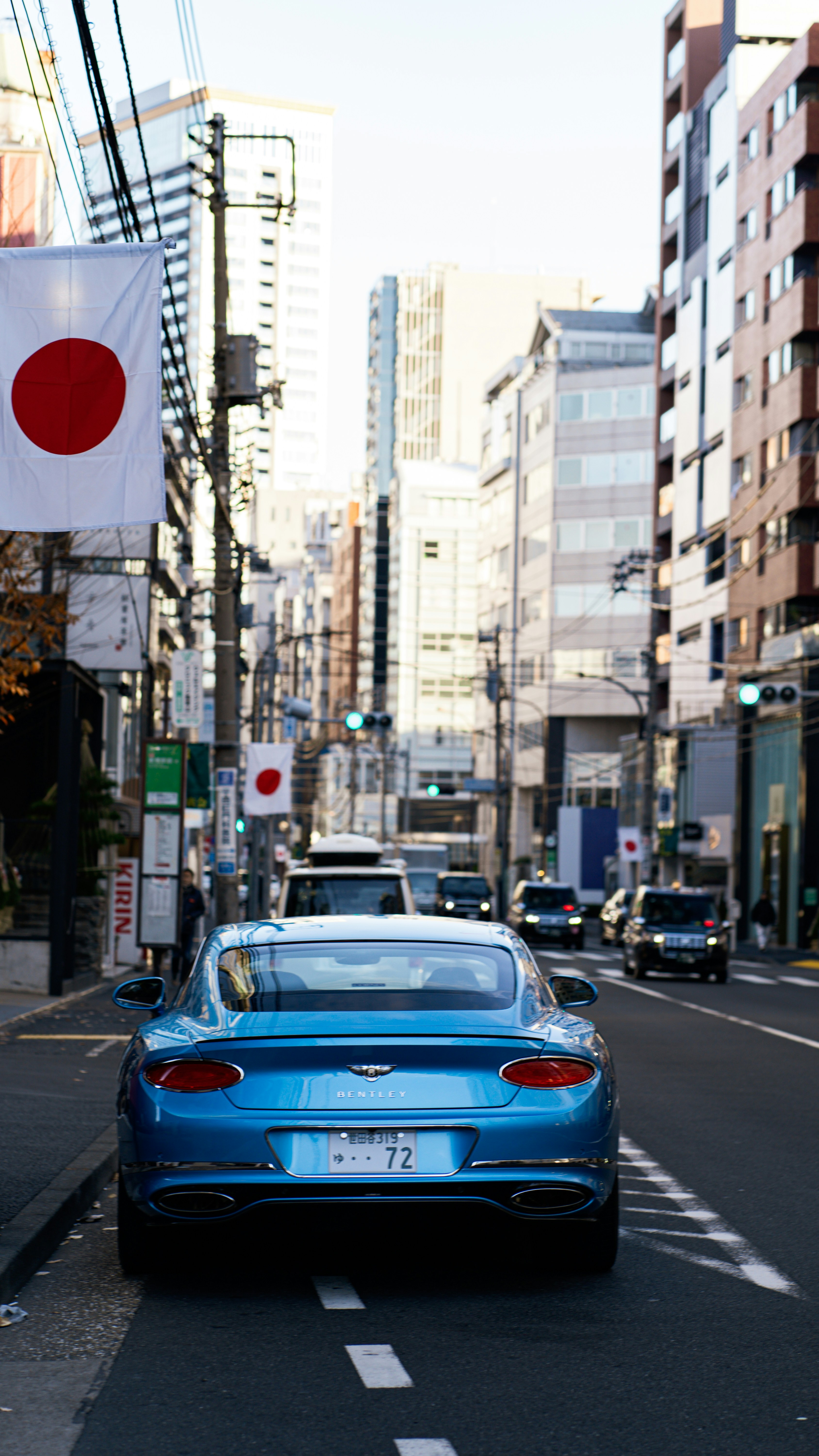 A striking blue Bentley parked along a downtown Tokyo street, with modern buildings and Japanese flags lining the road. This image blends high-end automotive luxury with urban Japanese aesthetics, capturing the quiet poise of a city moment.