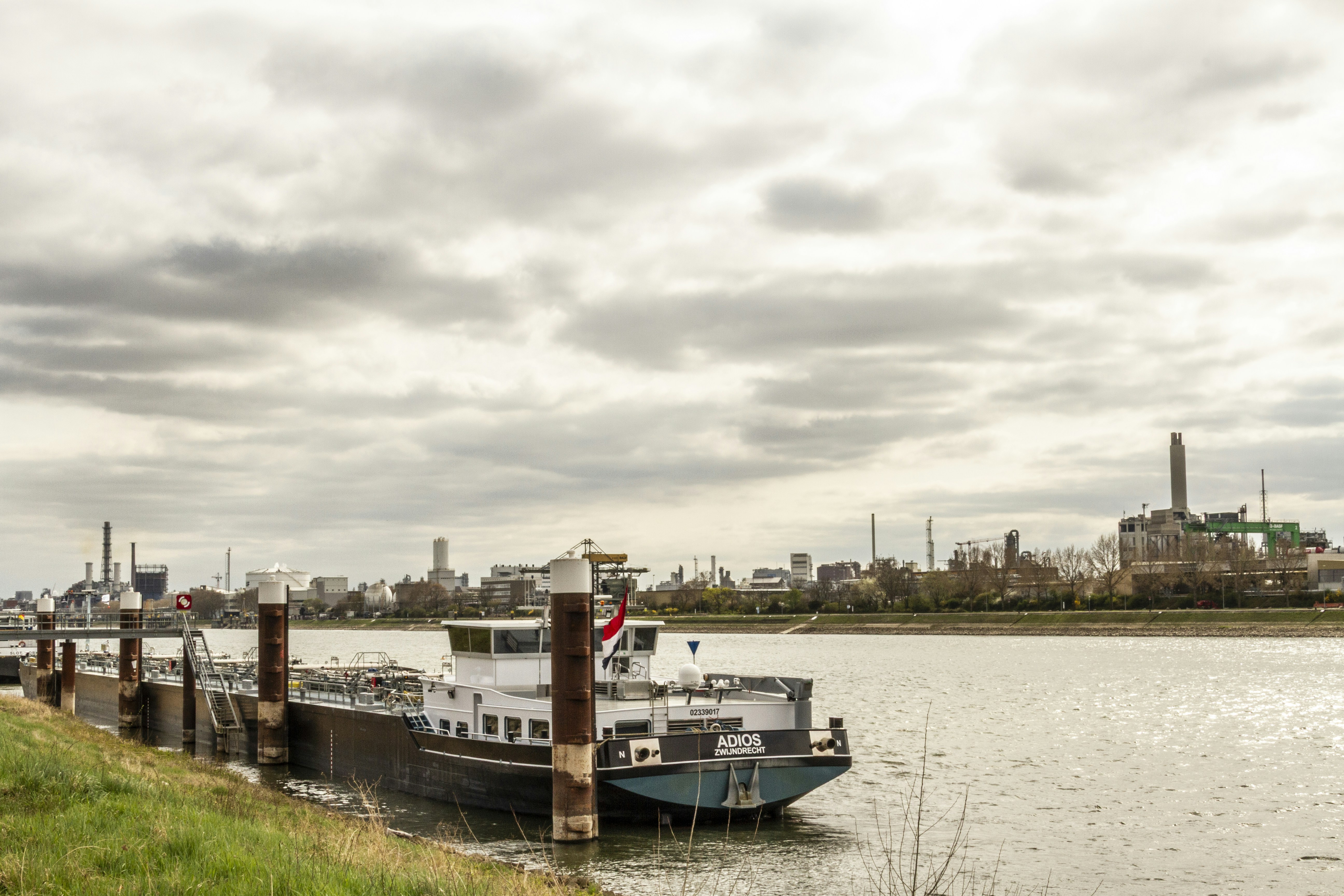 Boat moored at a riverside dock with industrial cityscape in the background under a cloudy sky.