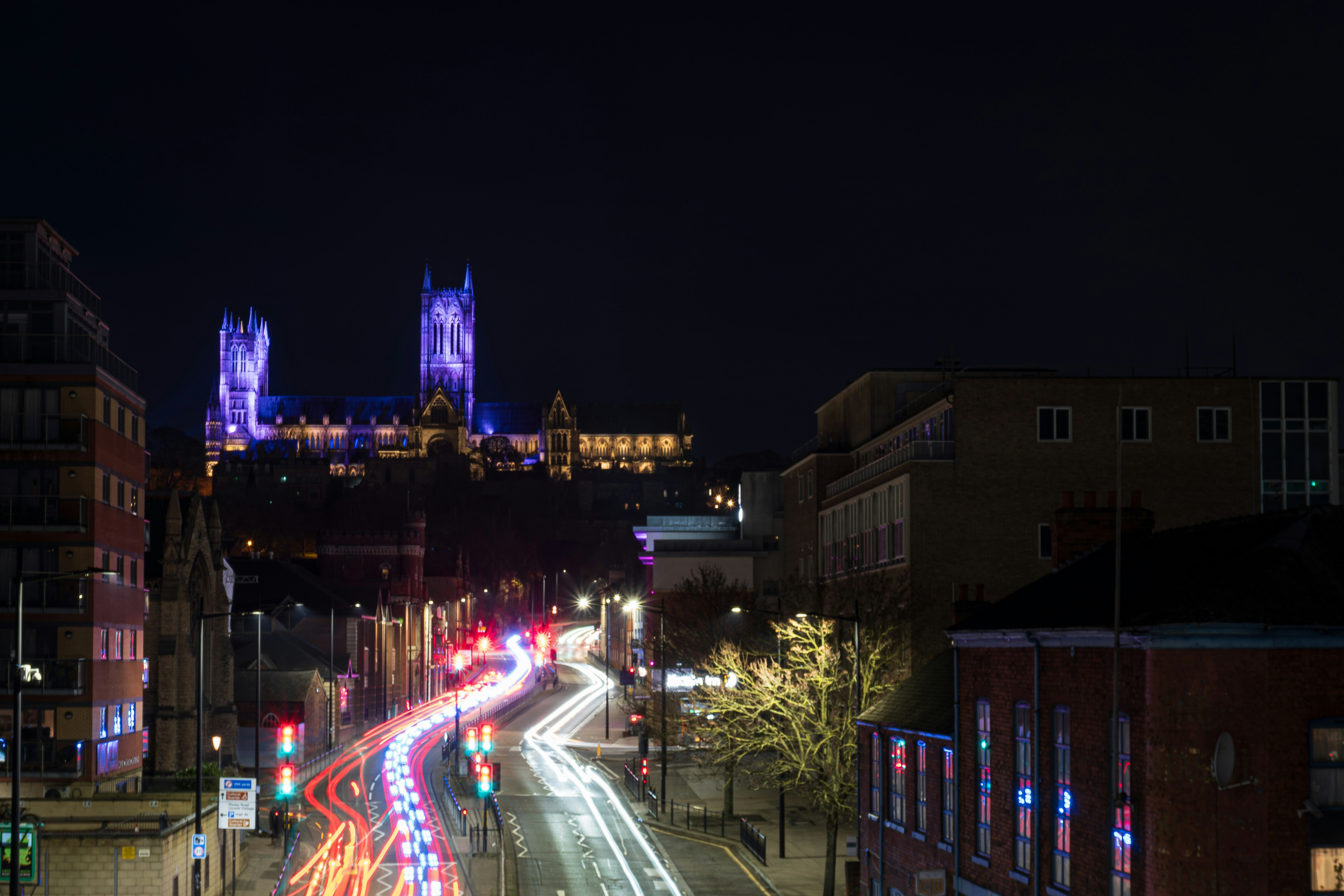 Historic cathedral illuminated in vibrant purple against a dark night sky, with streaks of car lights weaving through the urban landscape.