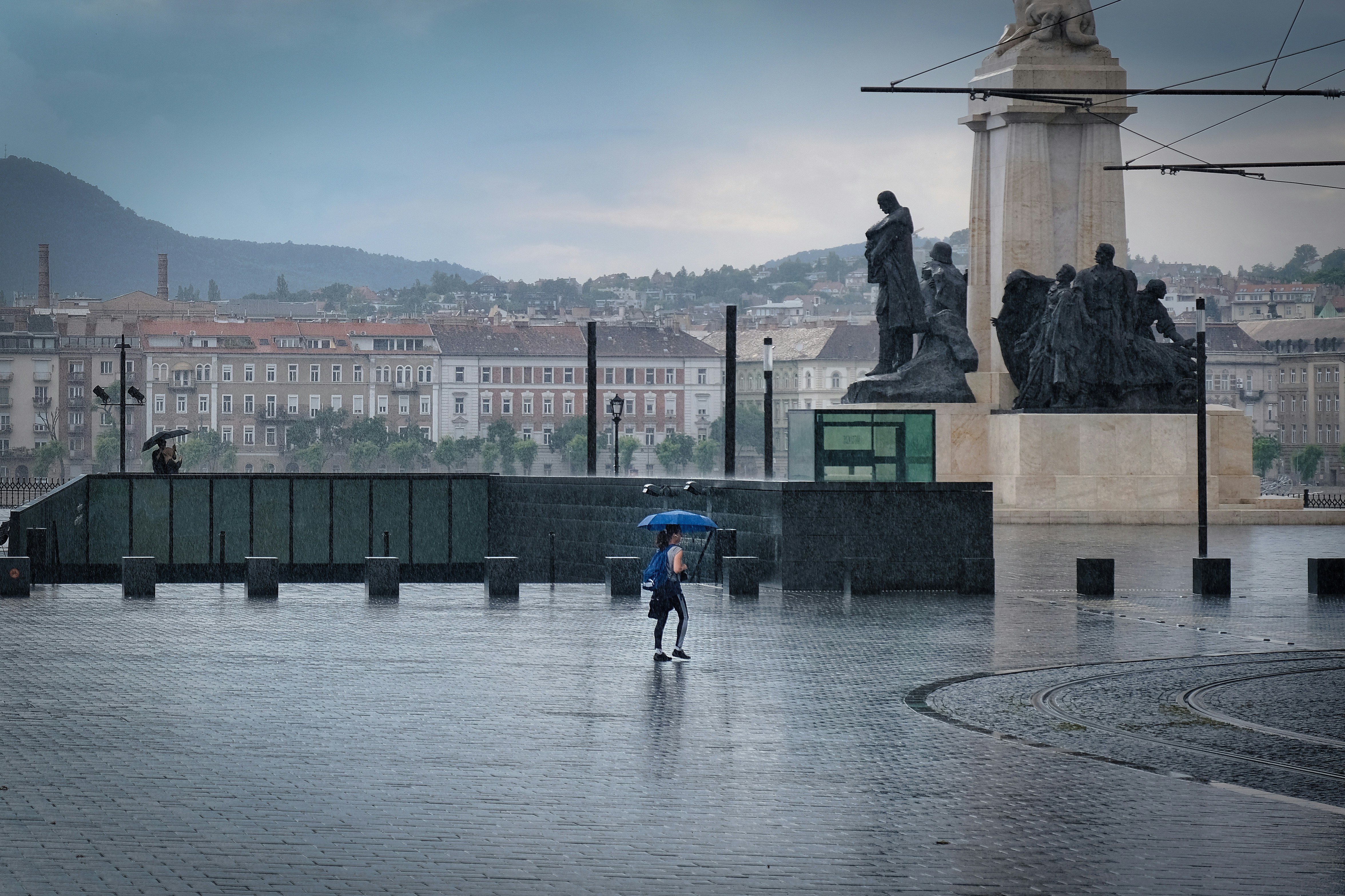 Person with a blue umbrella walks through a rain-soaked plaza, with a monument and cityscape in the background.