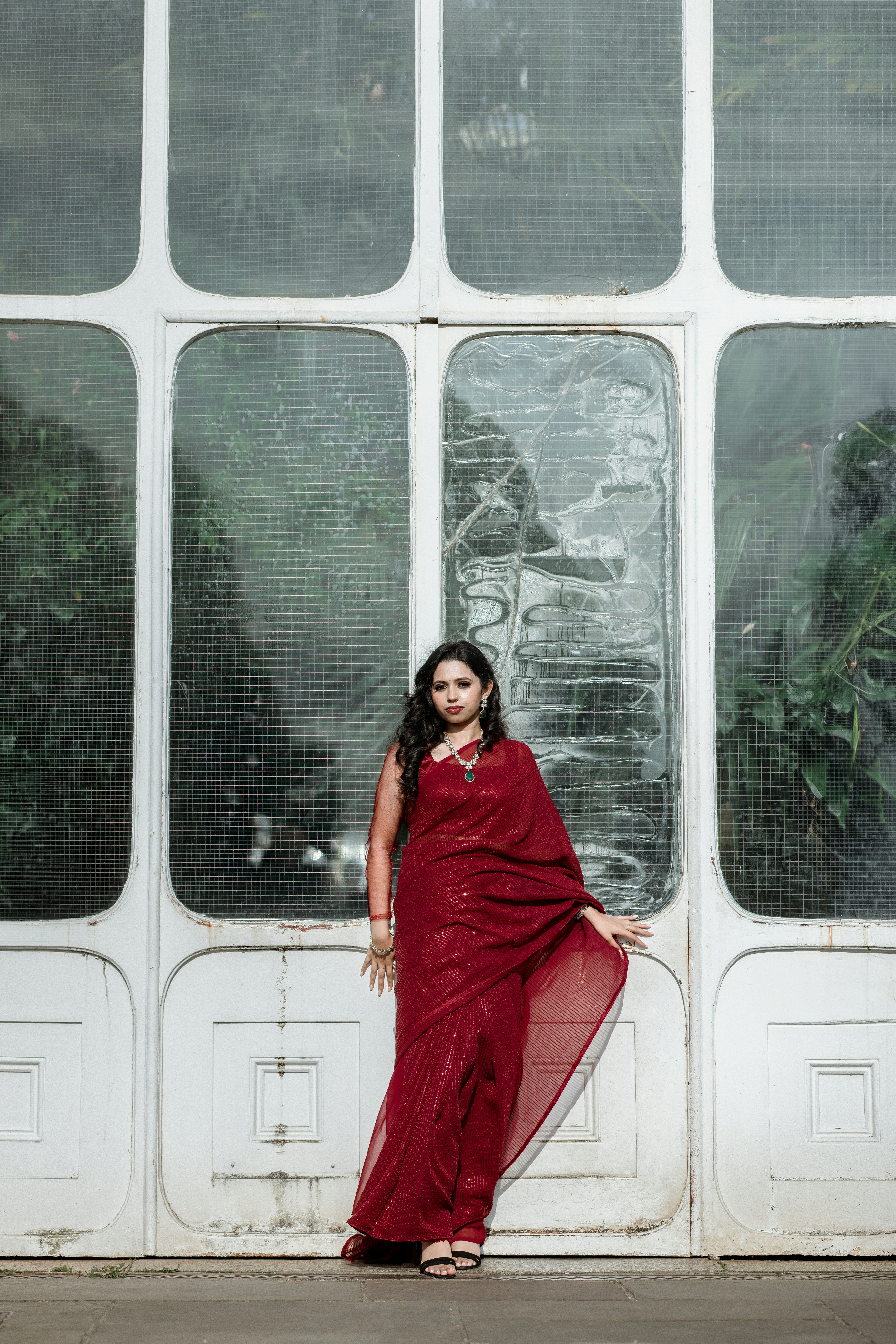 Woman in a red sari poses in front of large glass panels with lush greenery visible behind.