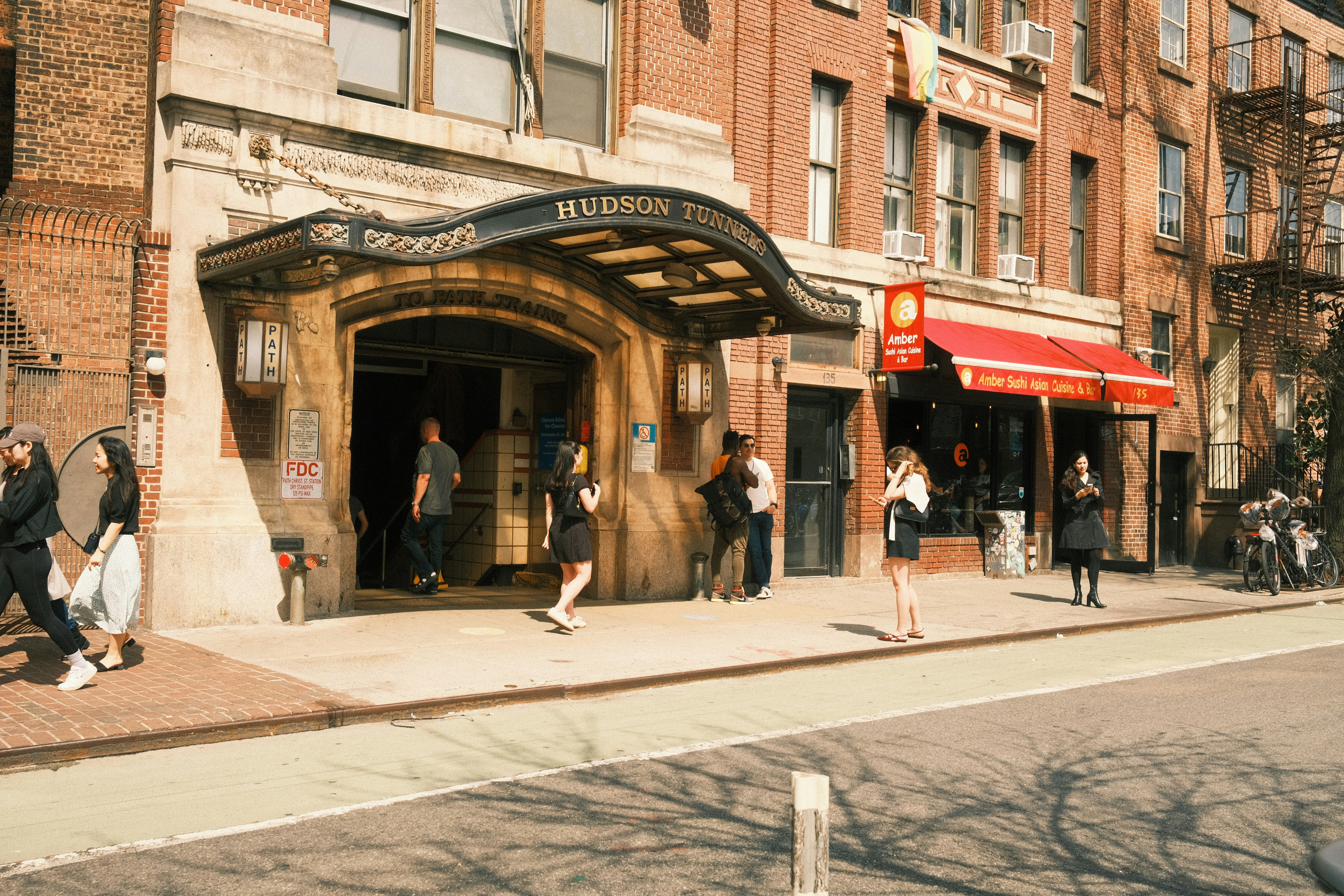 People walking past an ornate theater entrance and nearby café on a sunny street.