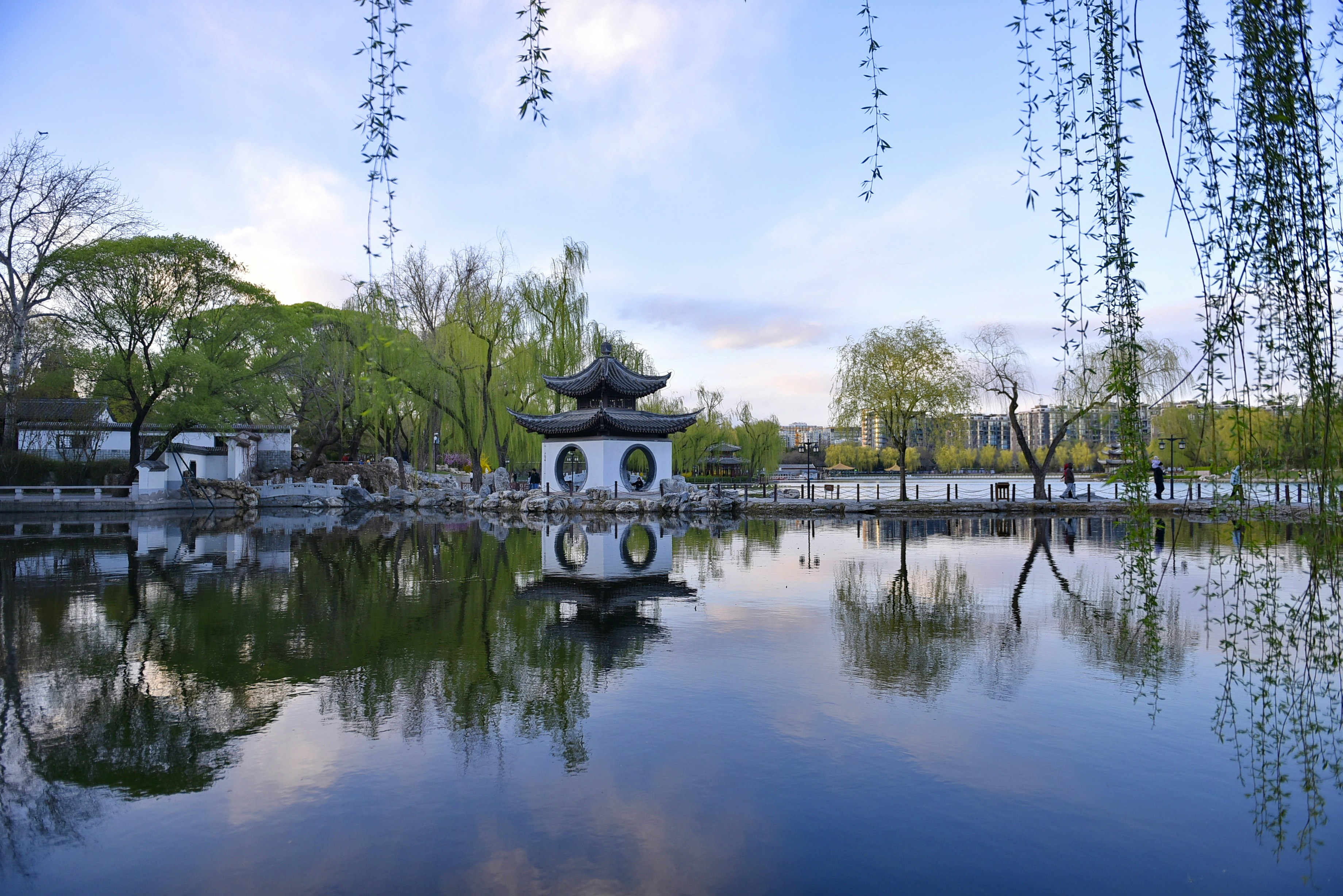 Serene pond with reflections of a traditional pavilion and lush trees under a clear blue sky.