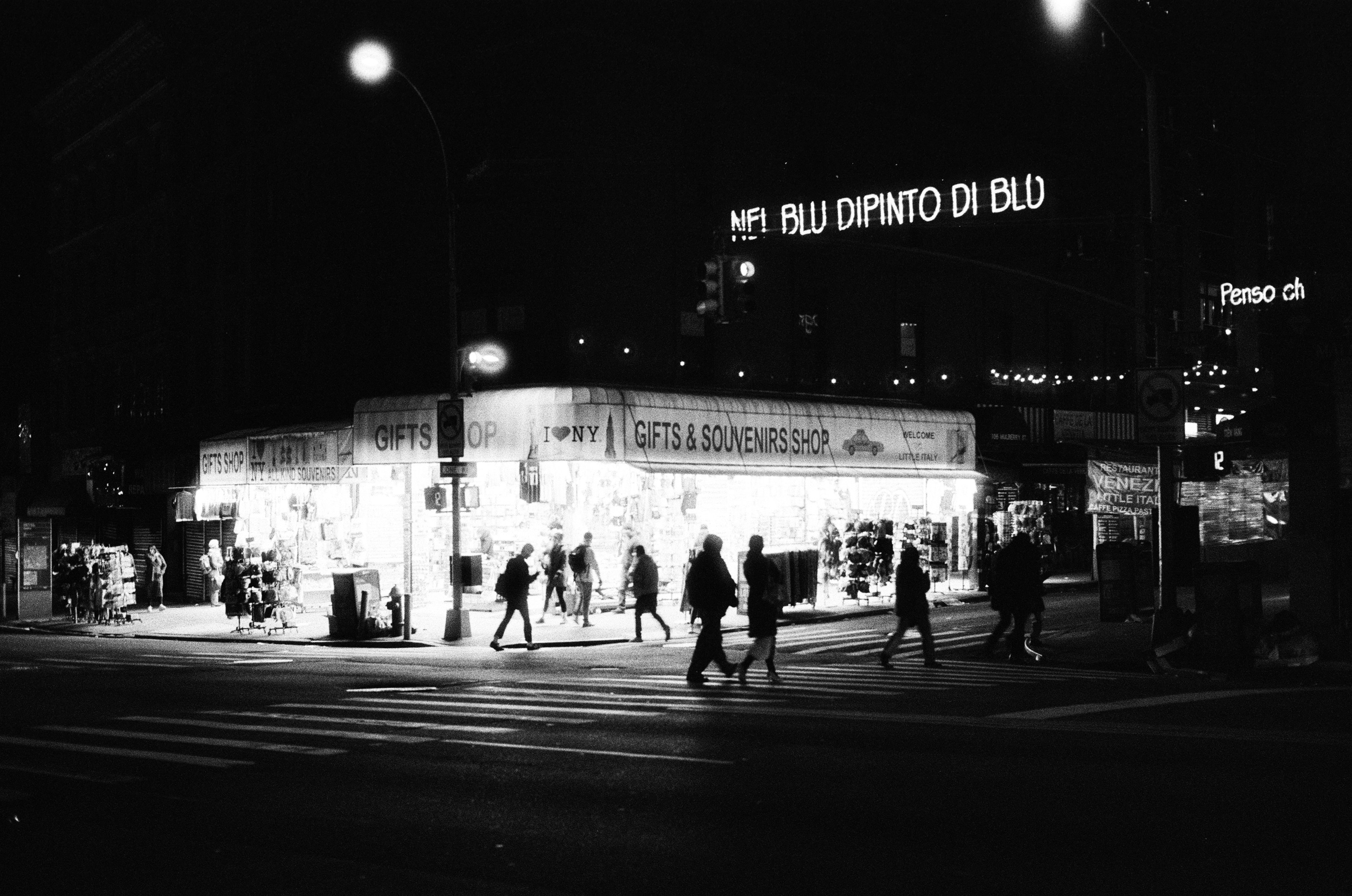 People cross a street at night, near illuminated storefronts. photo ...