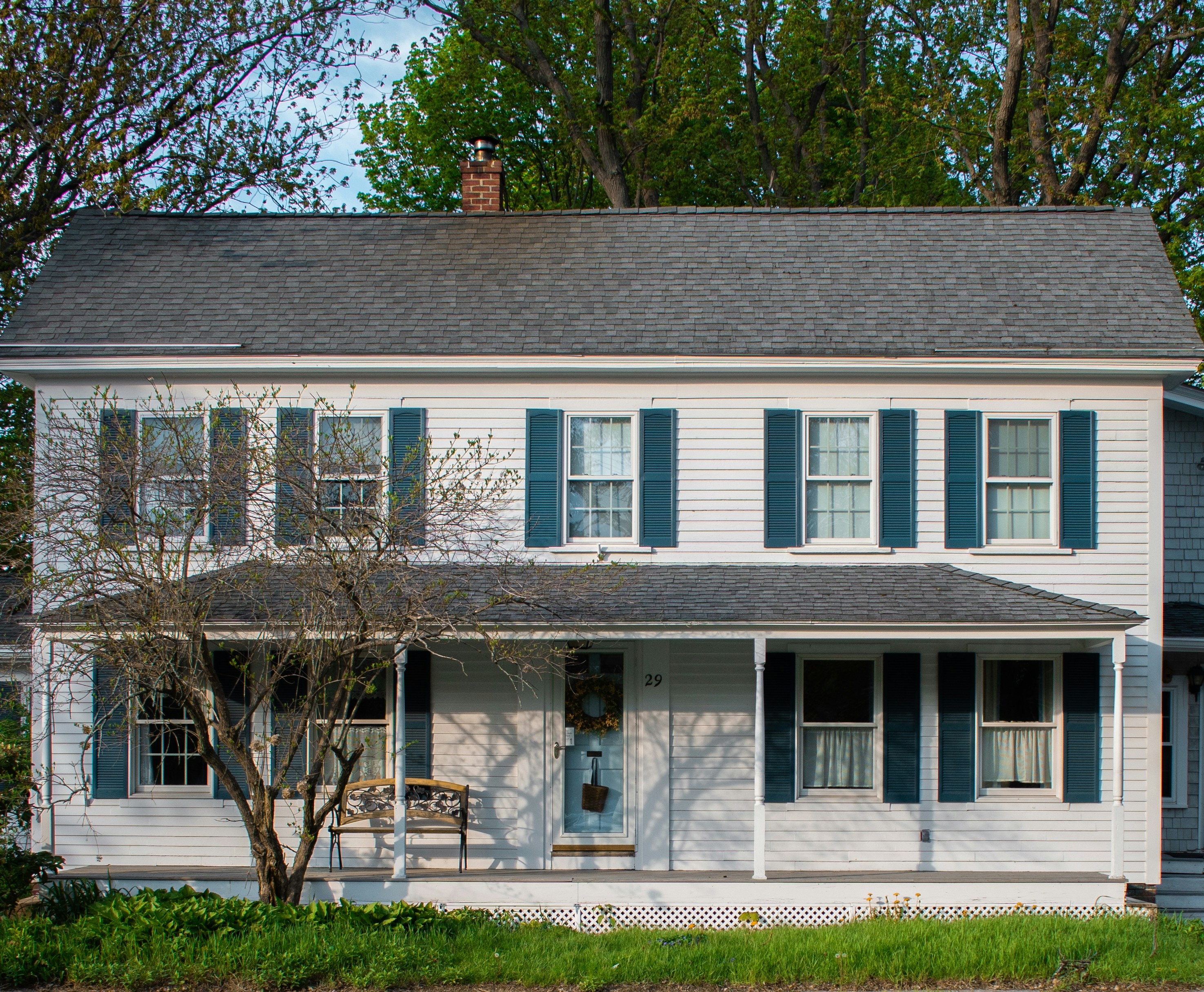 White colonial house with blue shutters and a front porch, surrounded by lush greenery under a clear sky.