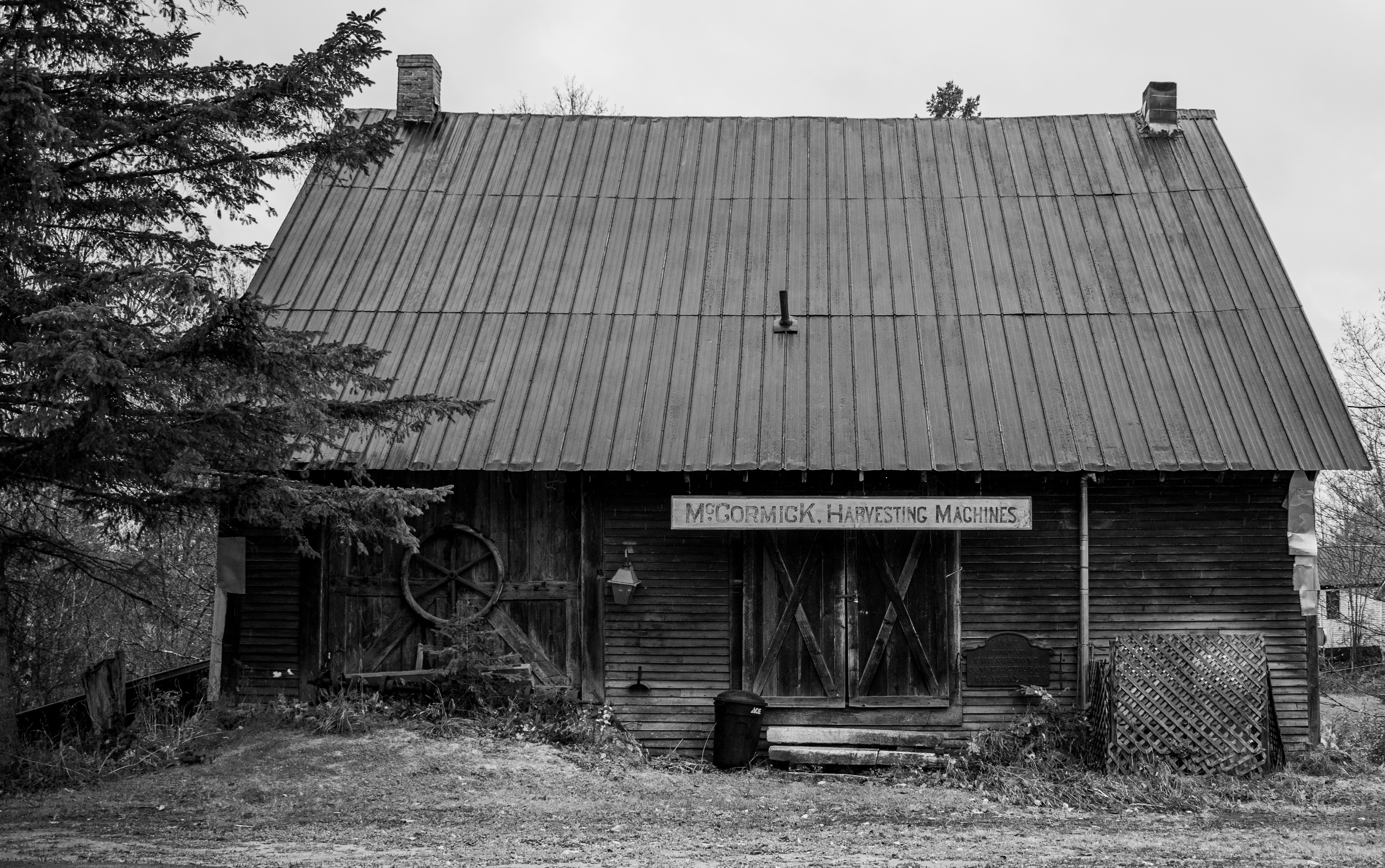 Black and white photograph of an old wooden house with a steep roof and boarded windows, surrounded by overgrown vegetation.