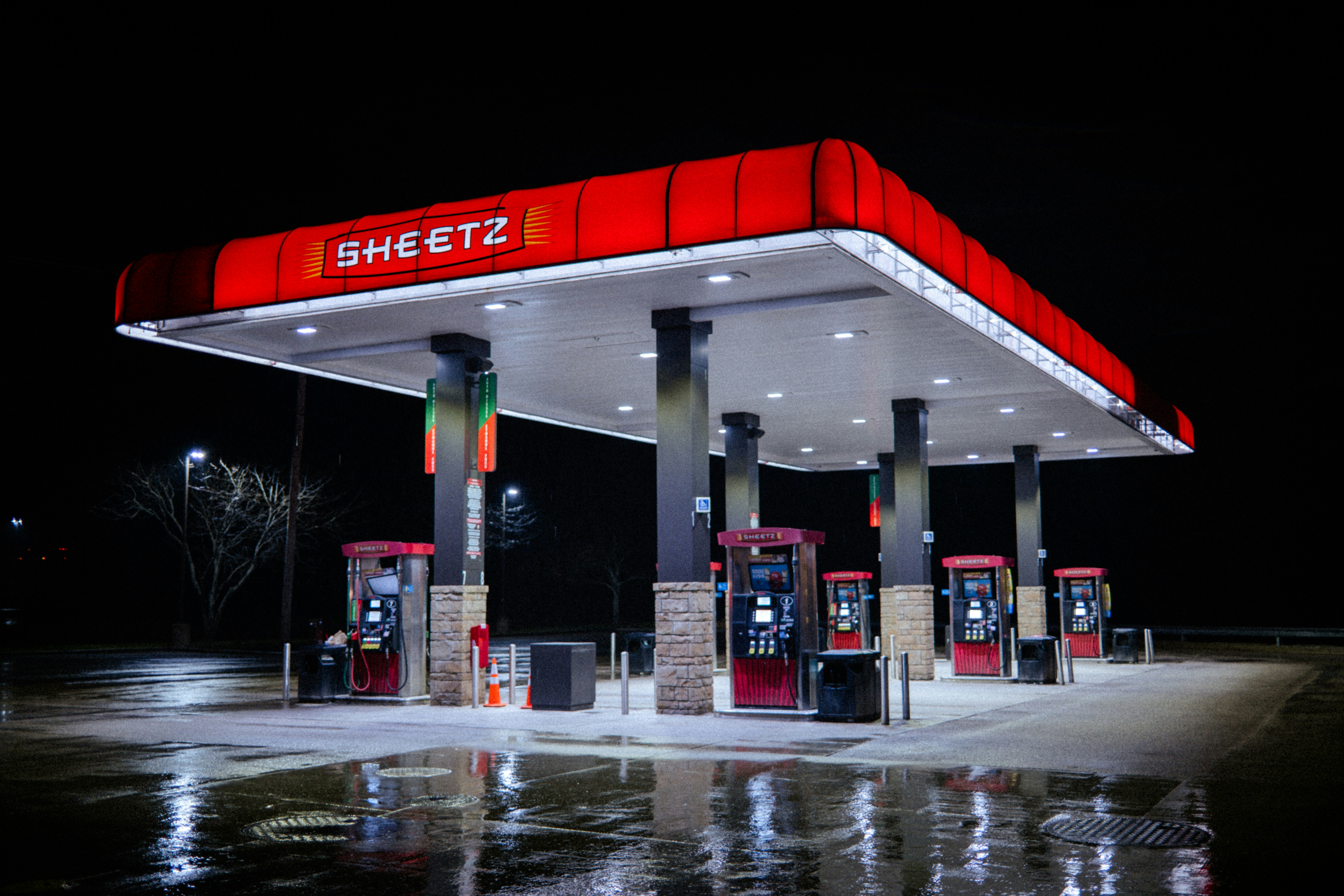 Illuminated gas station canopy casting reflections on wet pavement under a night sky.