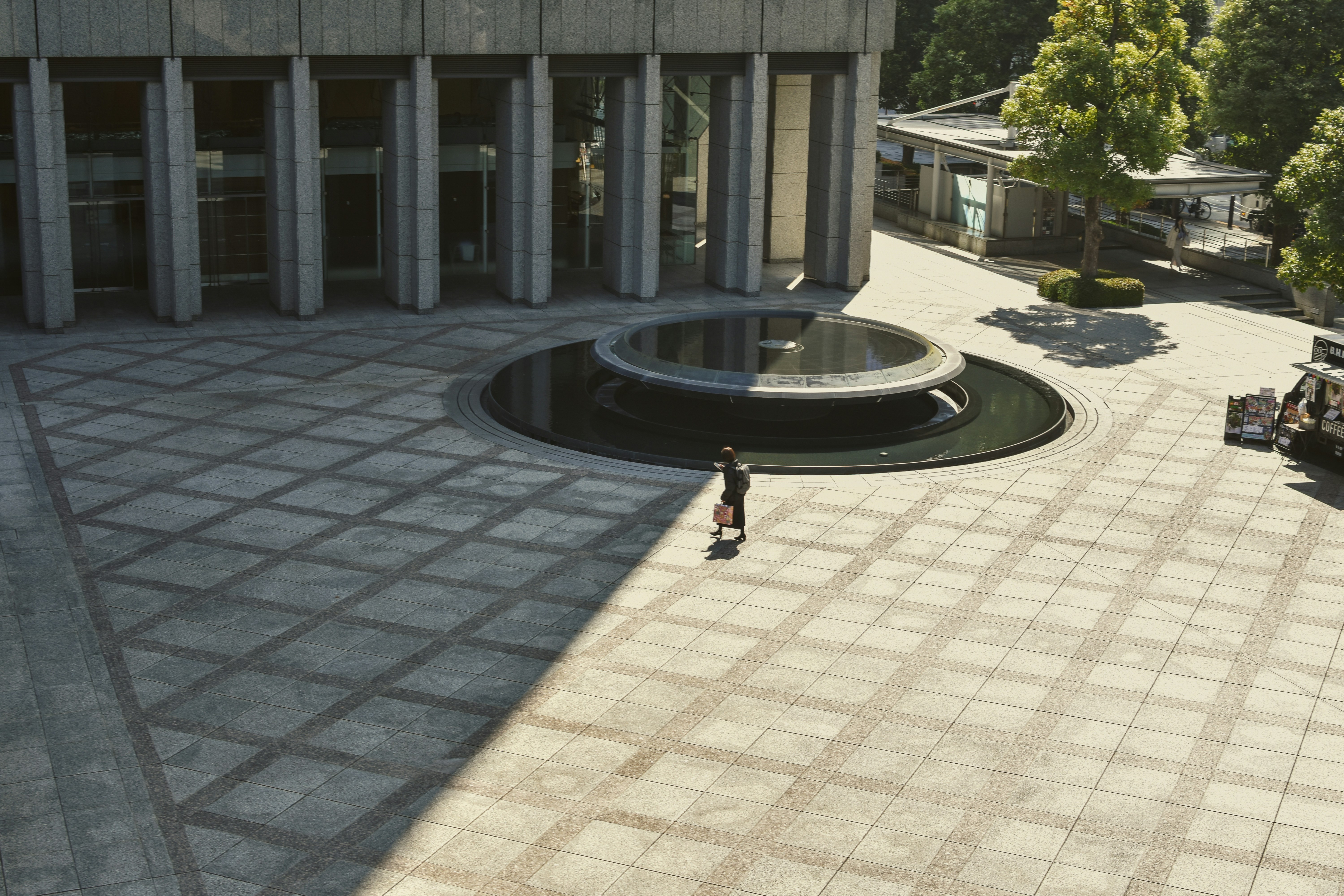 A lone figure stands on a patterned plaza, with a circular water feature reflecting the surrounding architecture. The interplay of light and shadow creates a dynamic atmosphere.