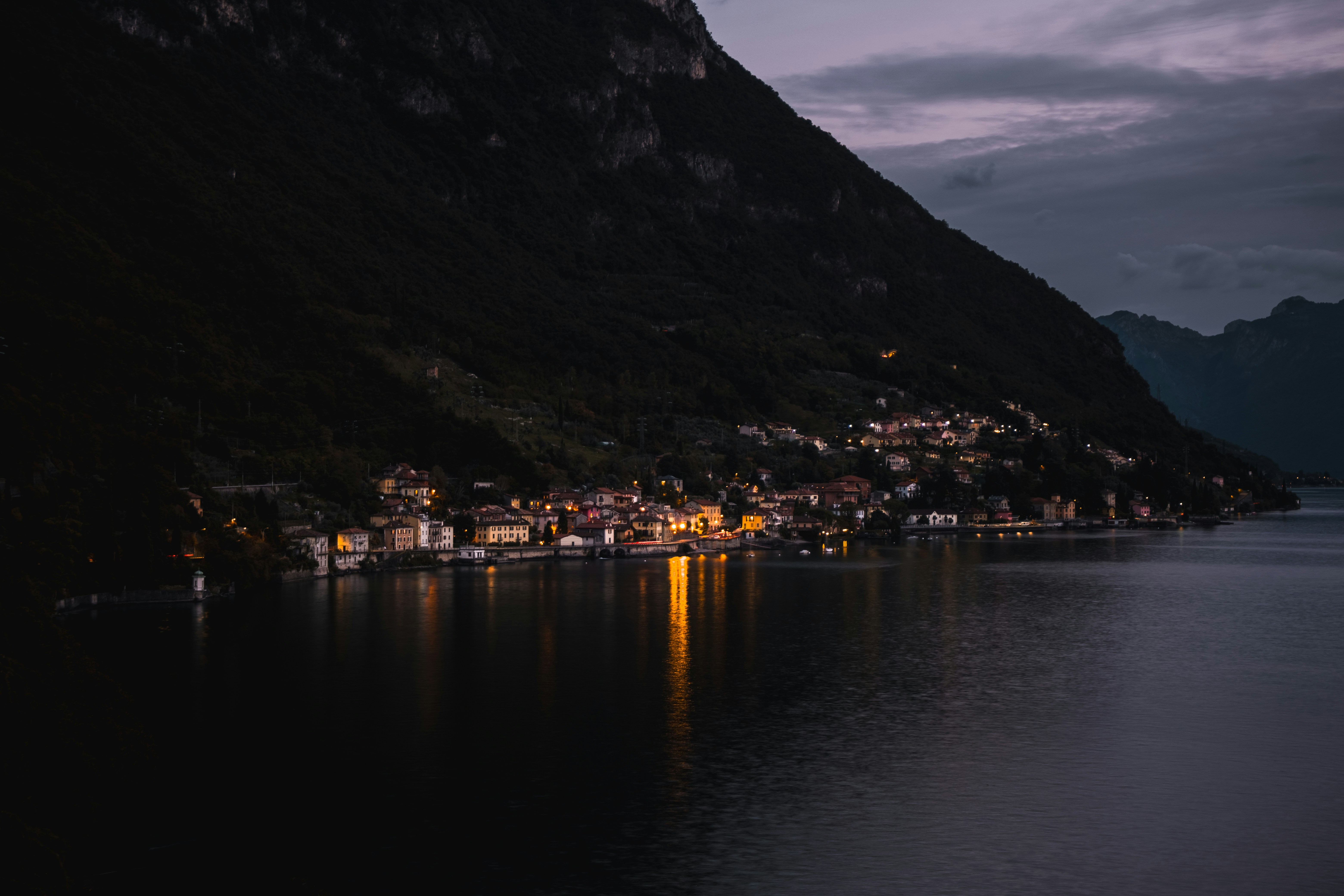 Evening lights reflect on a tranquil lake beneath a towering mountain under a dusky sky.