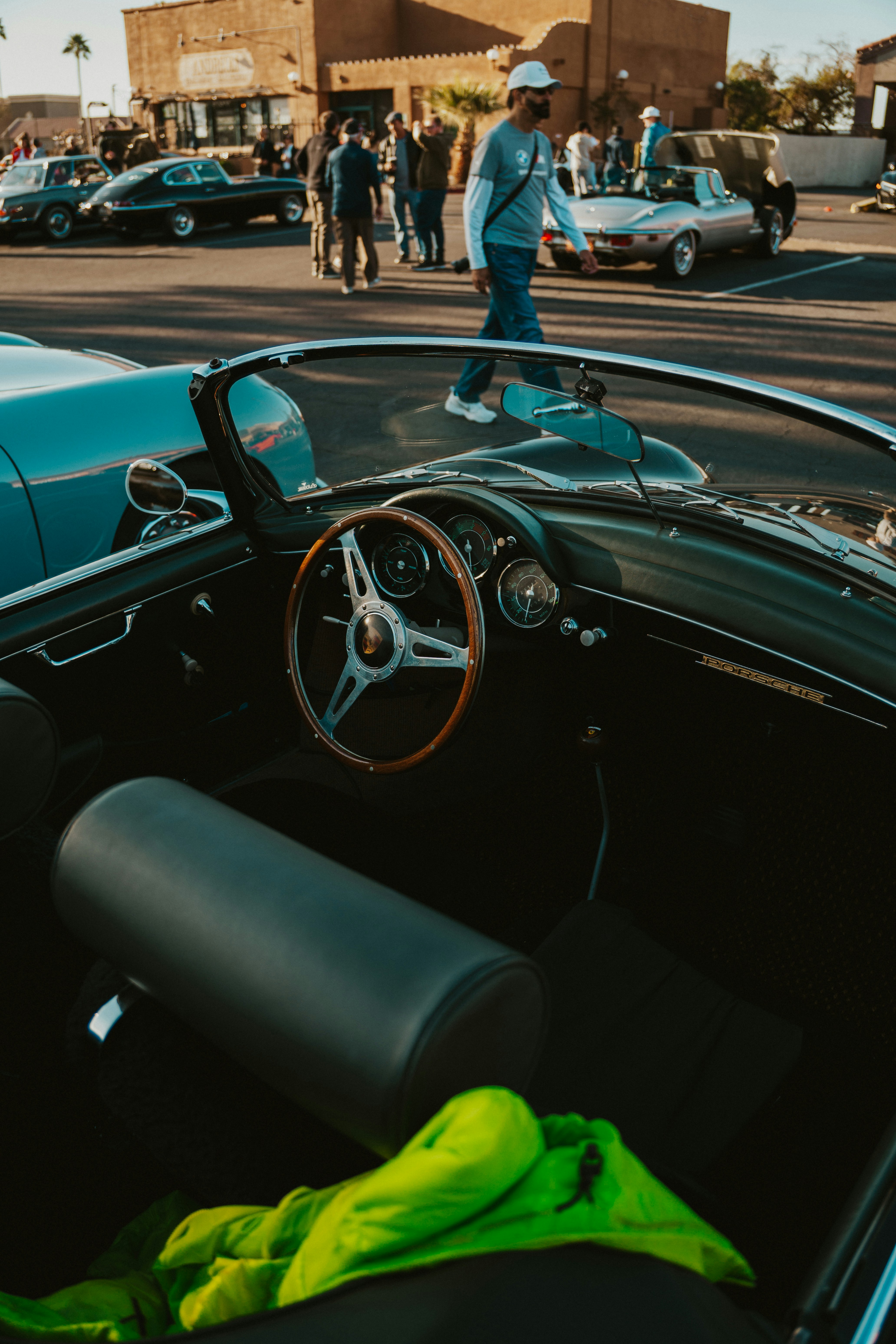 Interior view of a classic convertible car showcasing the steering wheel and dashboard, with a vibrant green jacket draped over the seat. The background features a gathering of vintage cars and enthusiasts.