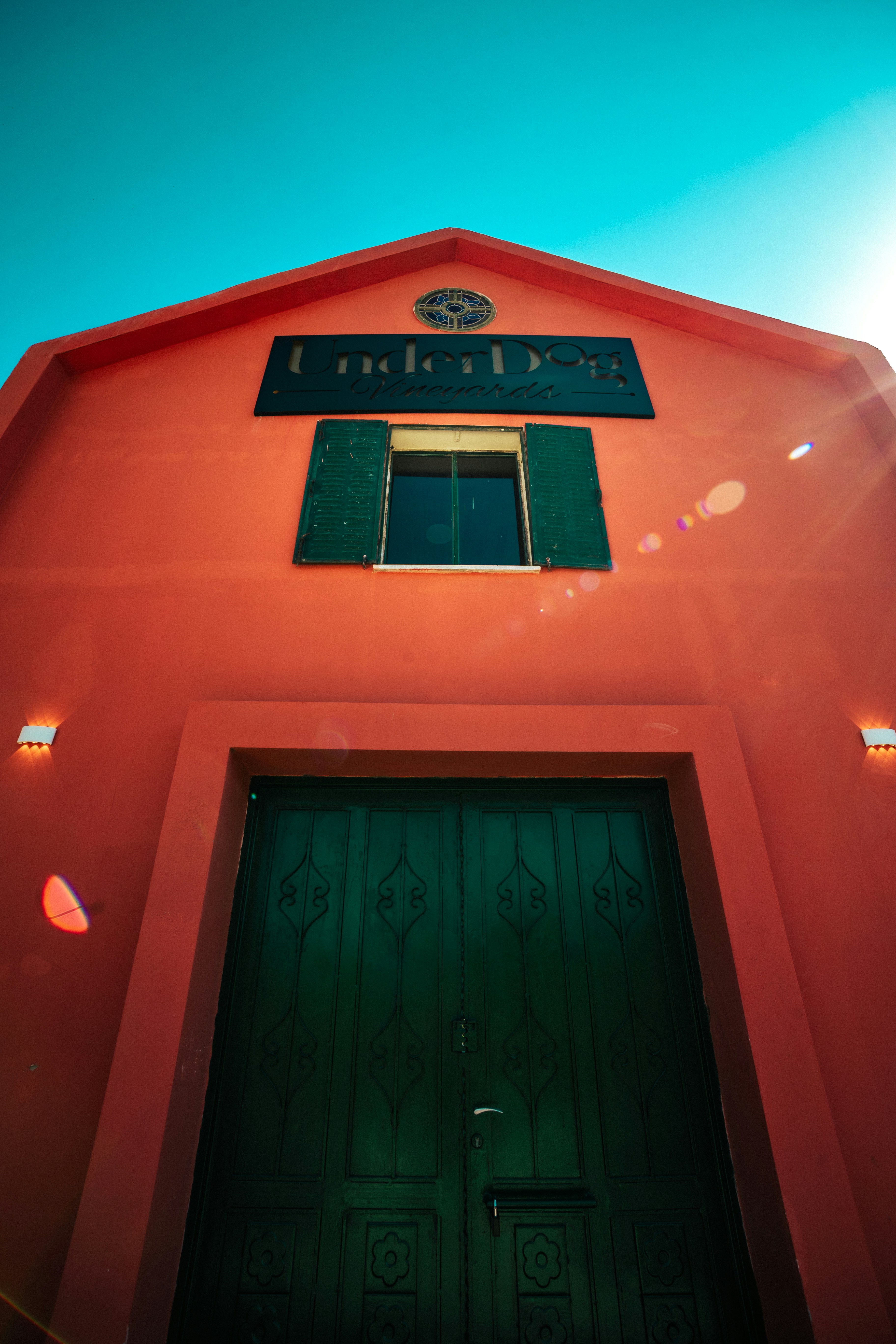 Vibrant orange building with a green door and shutters, illuminated by direct sunlight, creating lens flare effects.