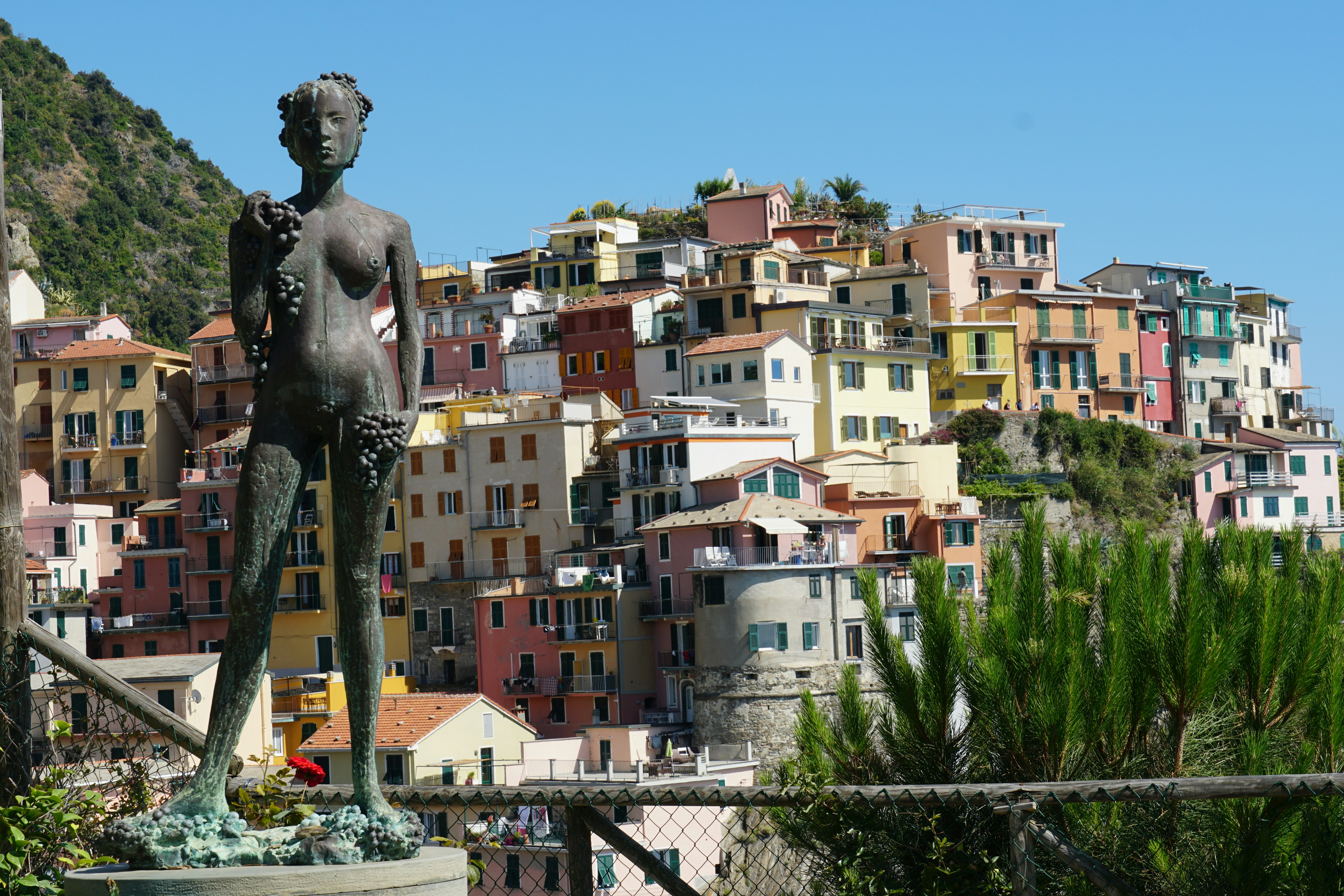 Bronze statue of a woman stands in the foreground, overlooking a vibrant hillside village with multicolored buildings stacked together.