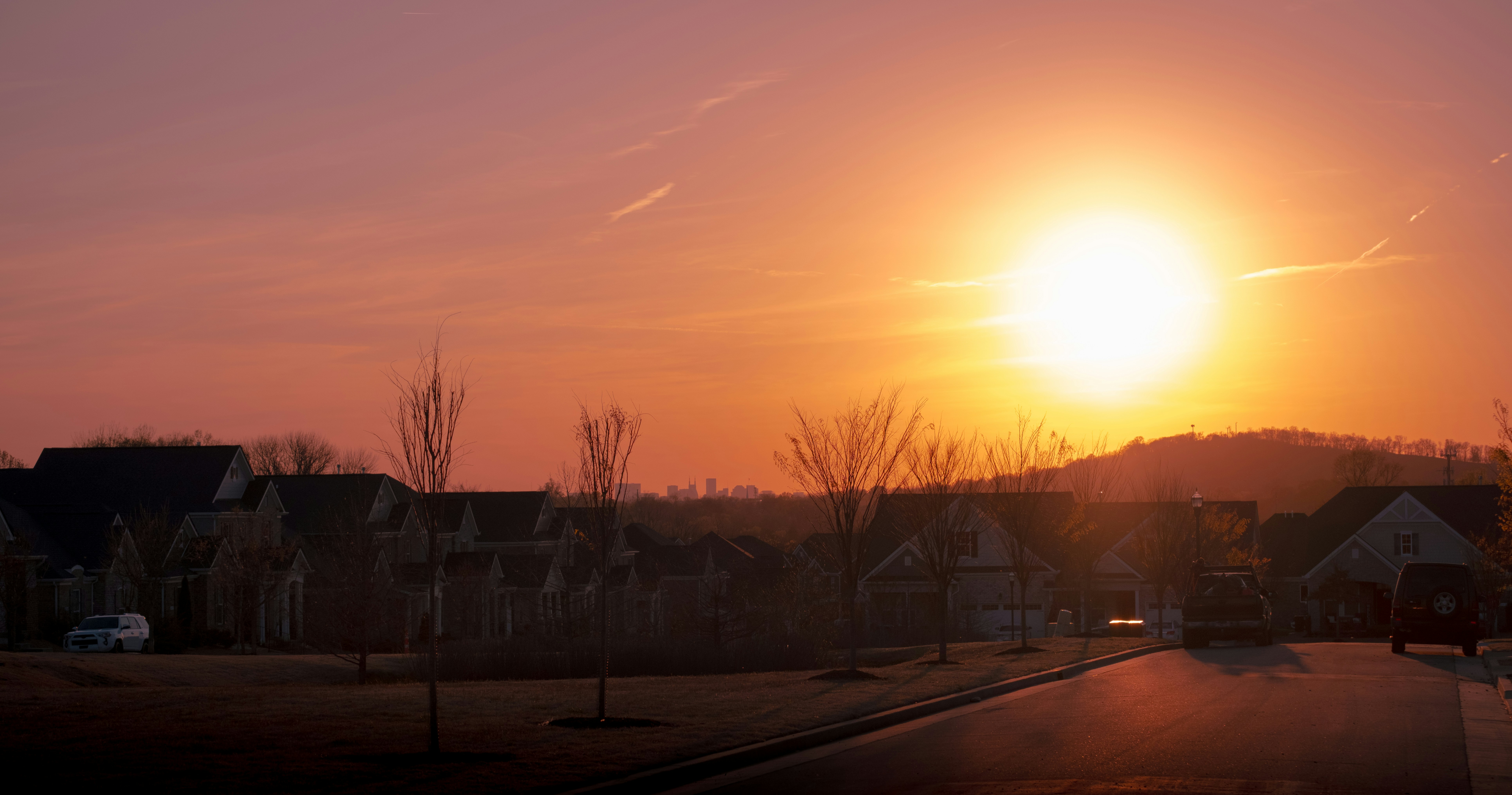 Sun setting over a suburban neighborhood with silhouetted houses and barren trees against a vibrant sky.