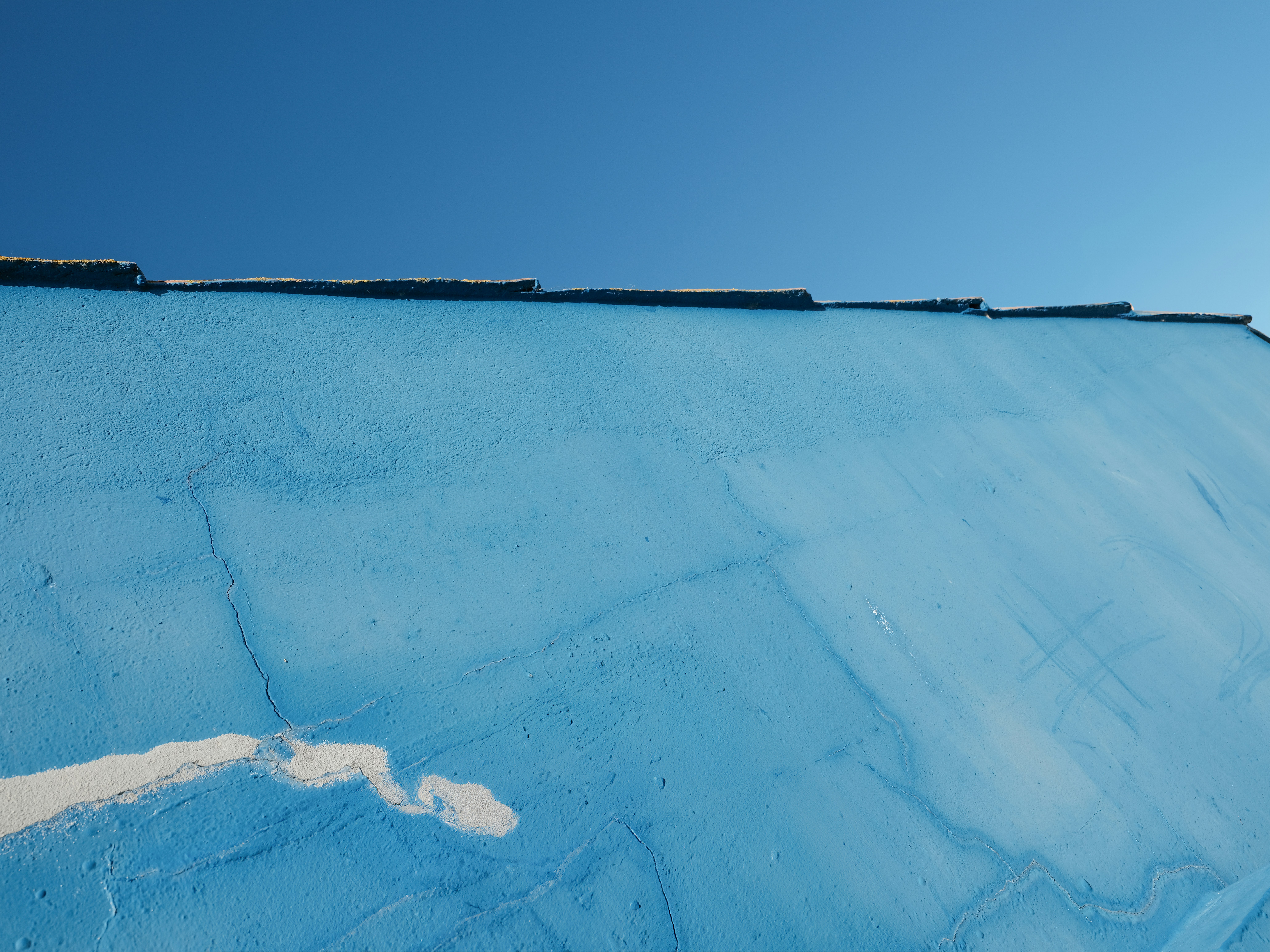 Photograph of a bright blue wall with a thin ridge along the top and a clear blue sky above.
