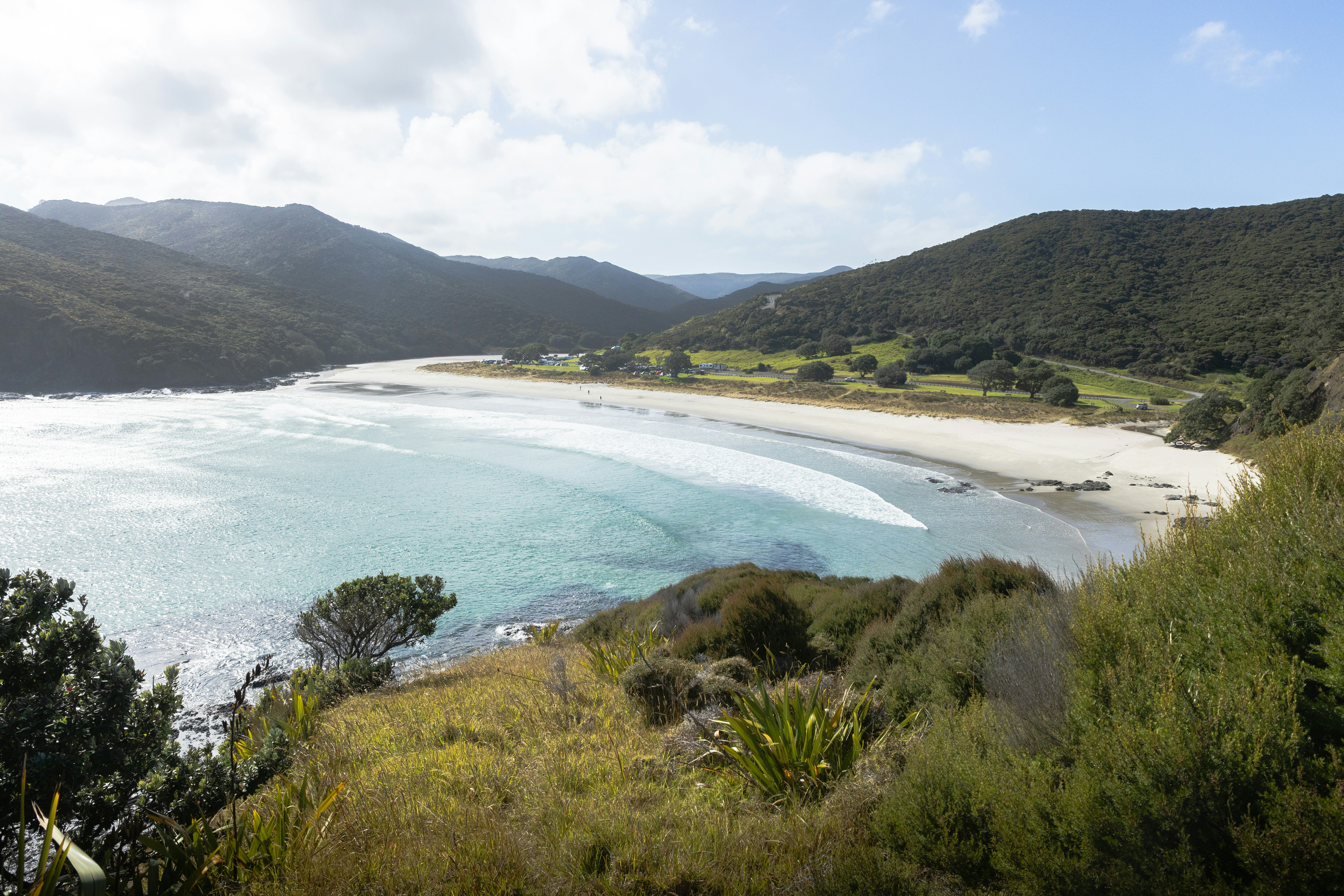 Expansive coastal landscape with gentle waves meeting a sandy beach, framed by lush green hills under a clear blue sky.
