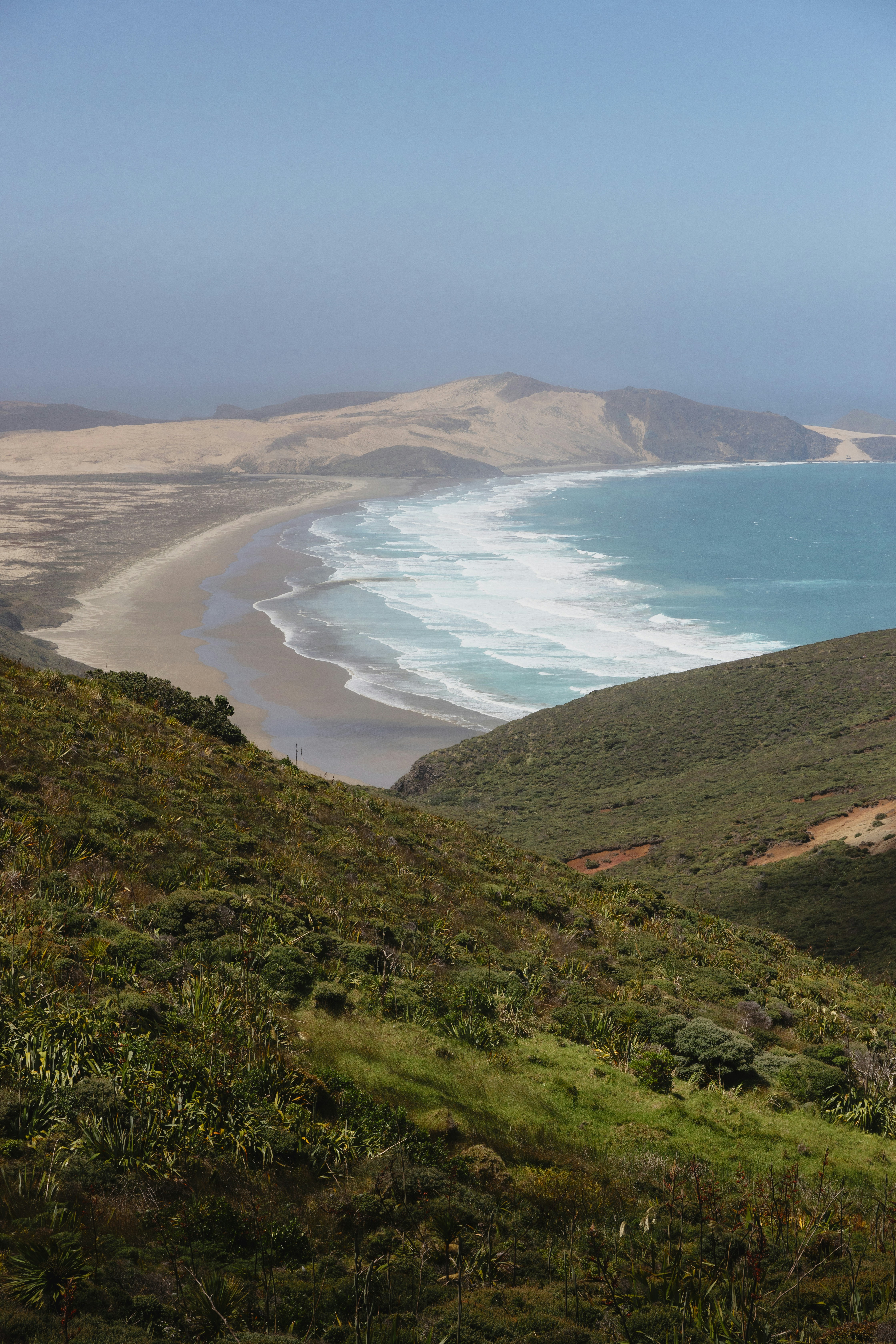 Sweeping coastline with rolling waves against sandy dunes and lush green hills under a clear blue sky.