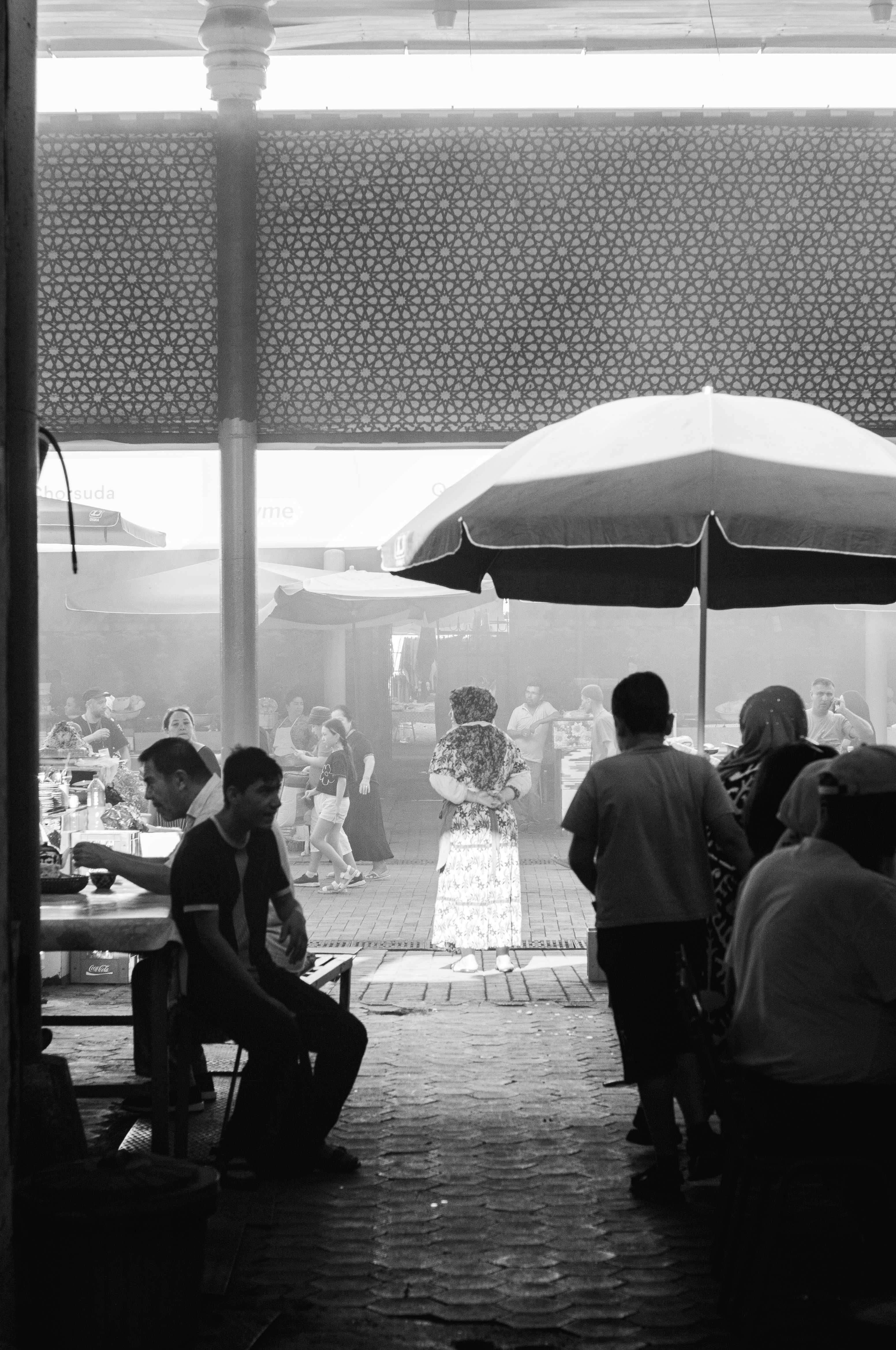 Monochrome street-café scene with a central figure in a light-patterned dress beneath an umbrella, surrounded by seated patrons and passing pedestrians. A candid photograph capturing everyday life in a busy plaza.