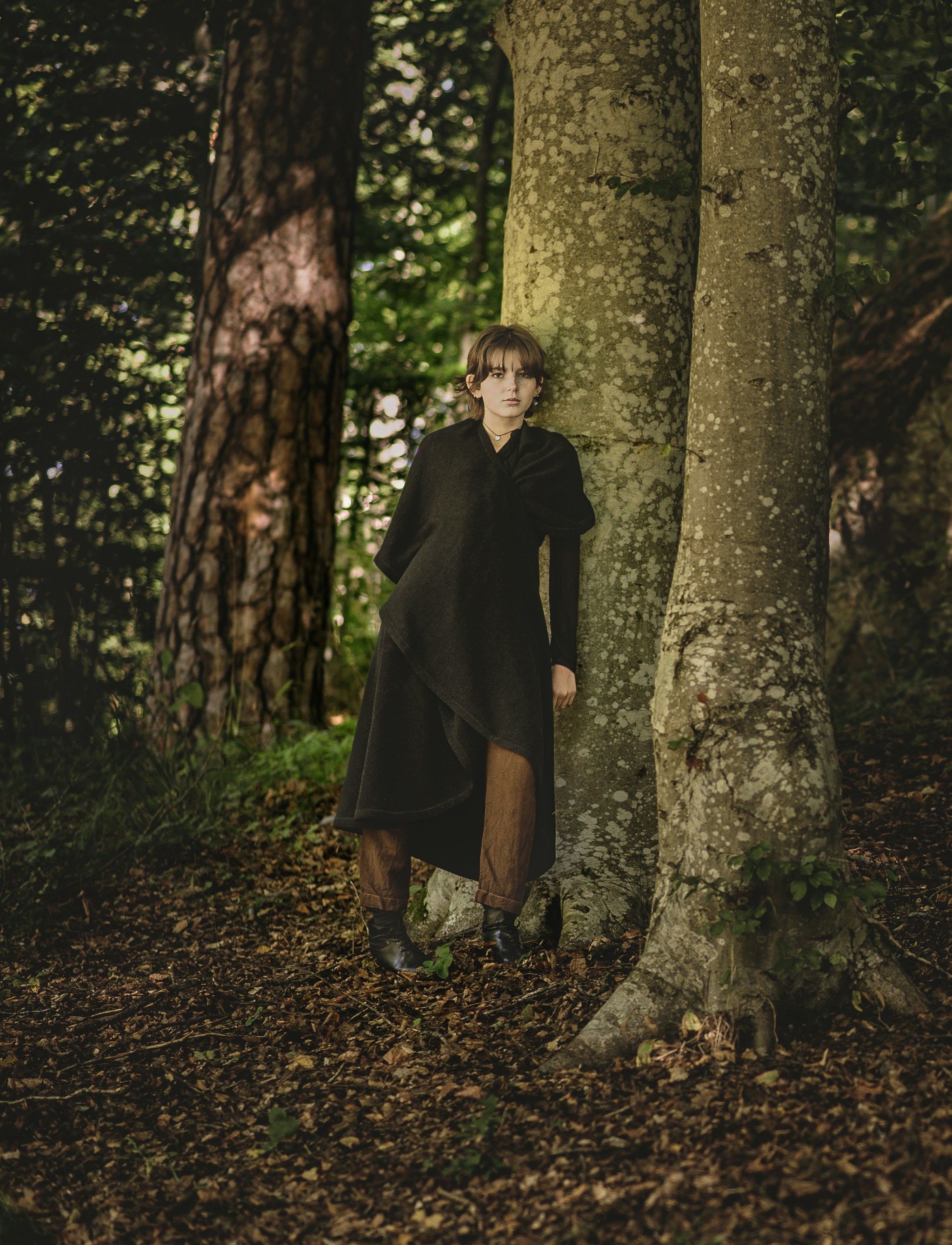 Person in dark clothing stands against large tree trunks in a forest setting.