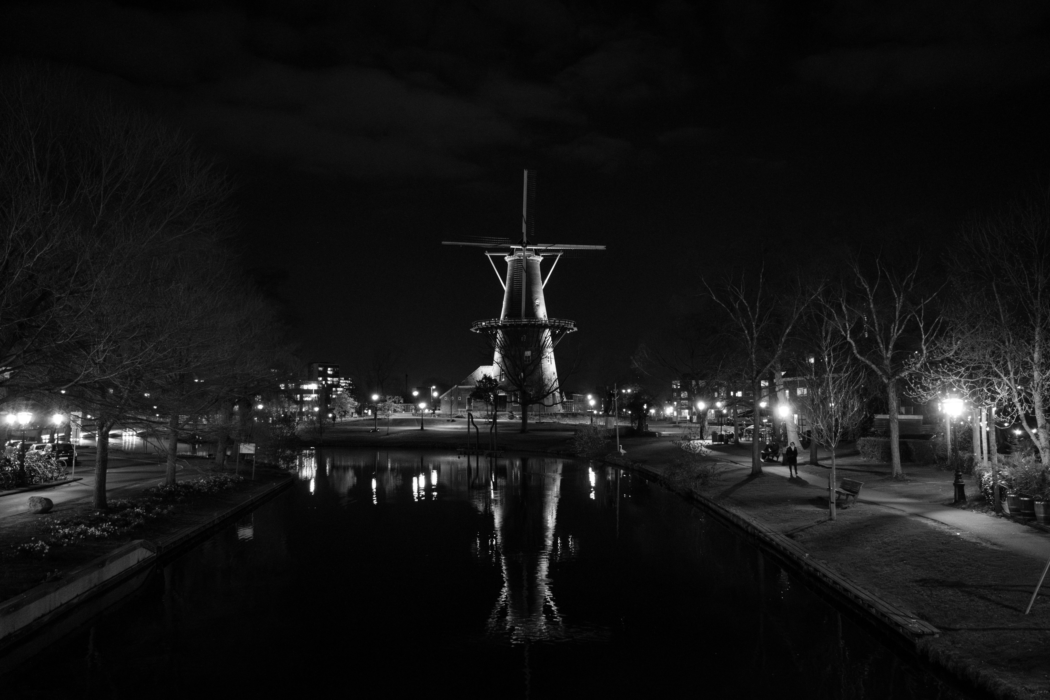 Black and white photo of a windmill reflected in a canal at night, surrounded by softly lit trees and streetlights.