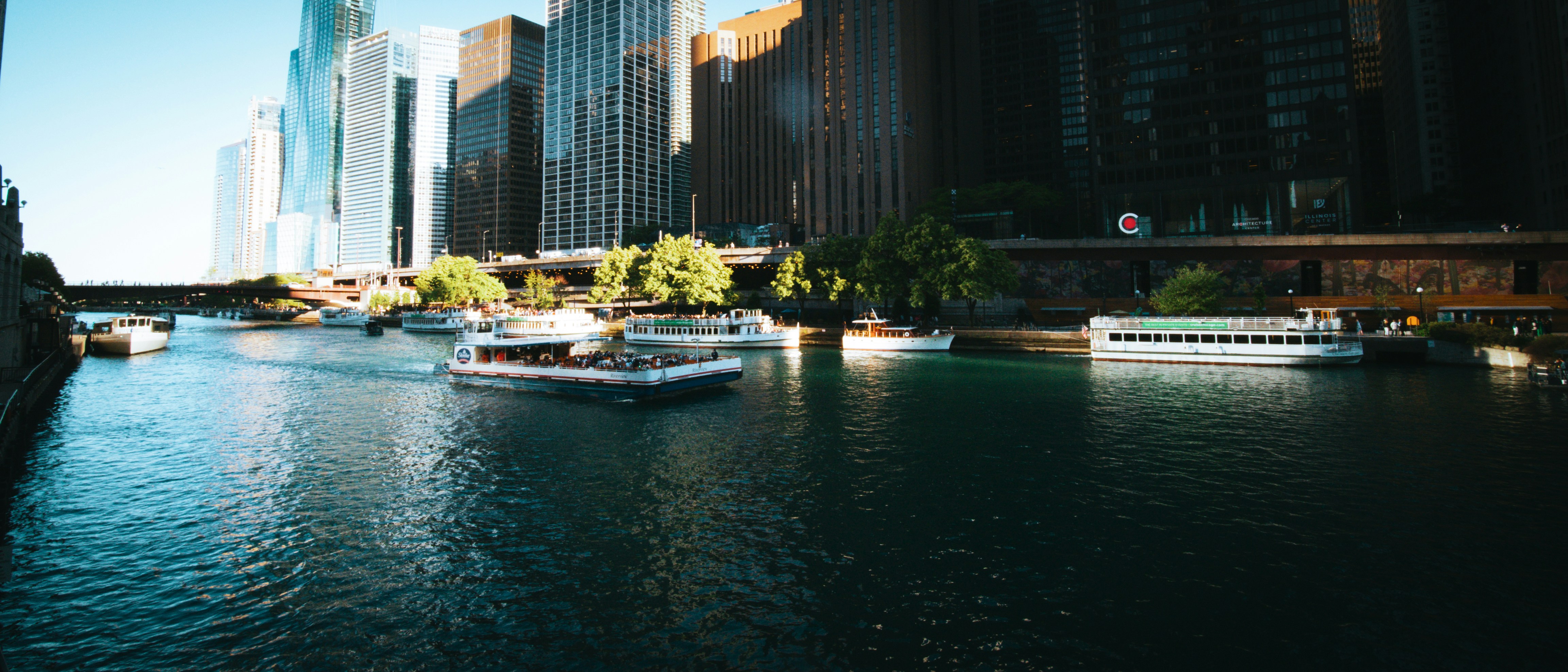 Boats navigating a sunlit river flanked by towering city skyscrapers.