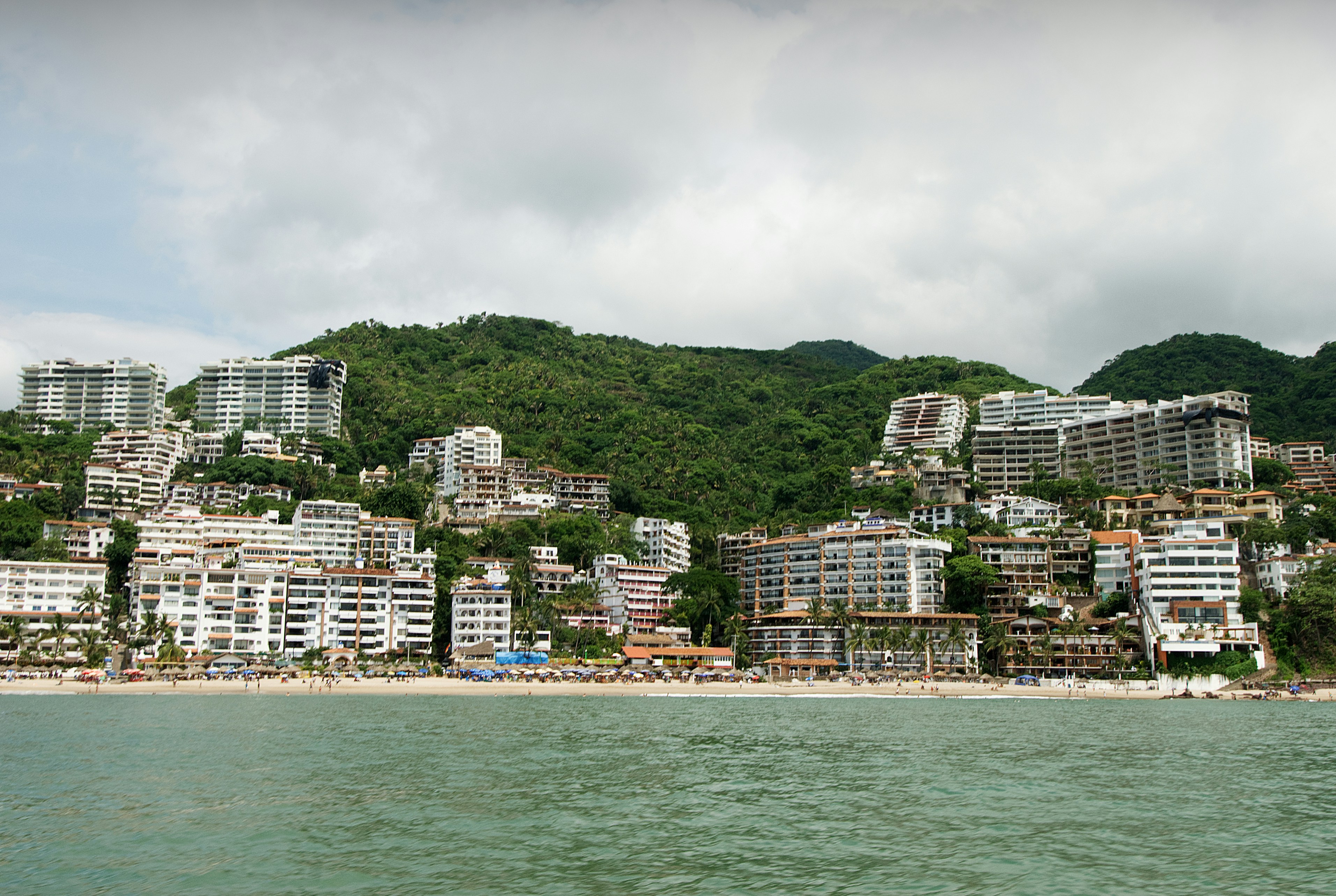 Coastal cityscape with buildings nestled against lush green hills under a cloudy sky.