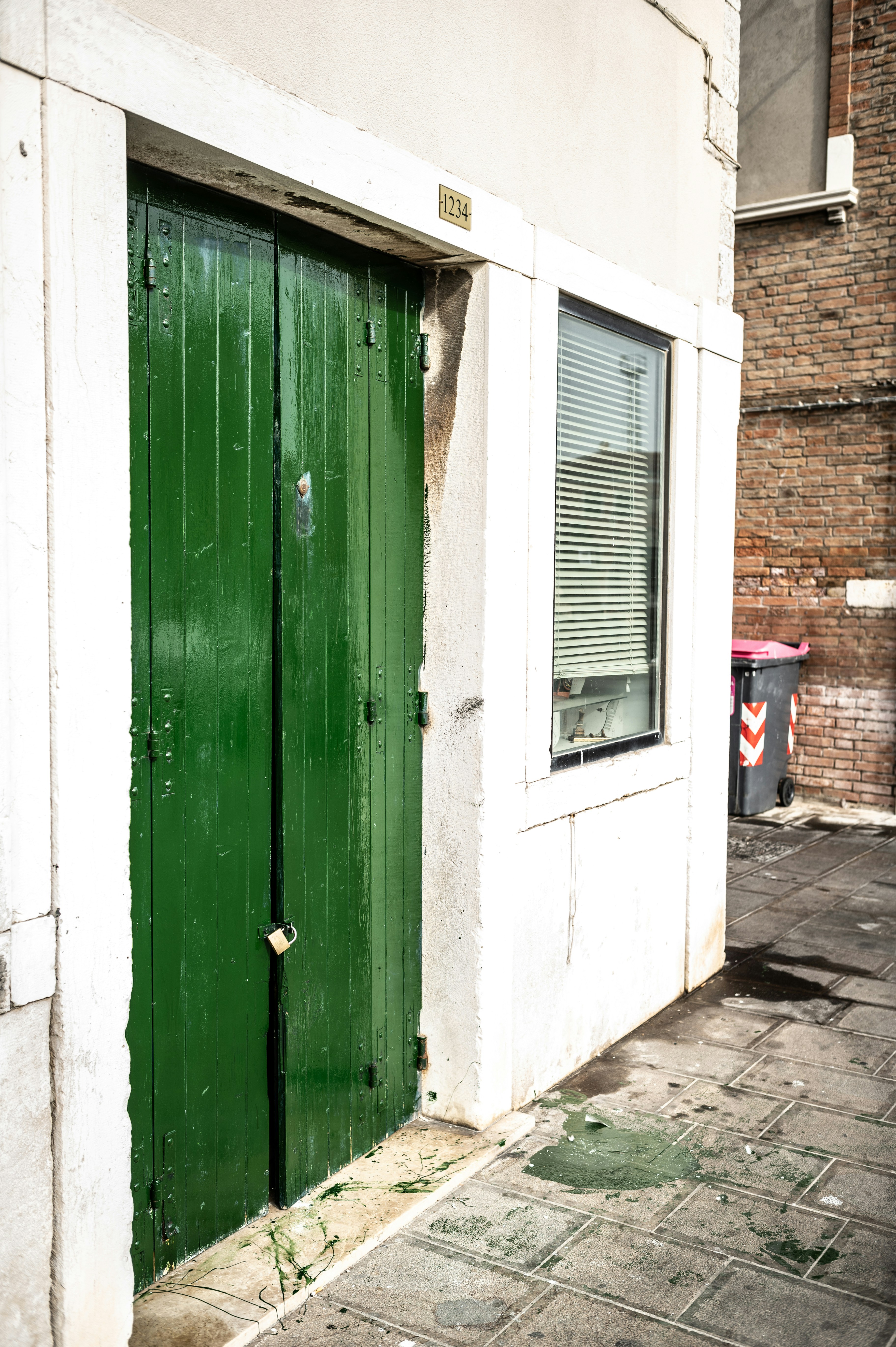 Green wooden door next to a window on a narrow street corner, with a hint of urban wear.