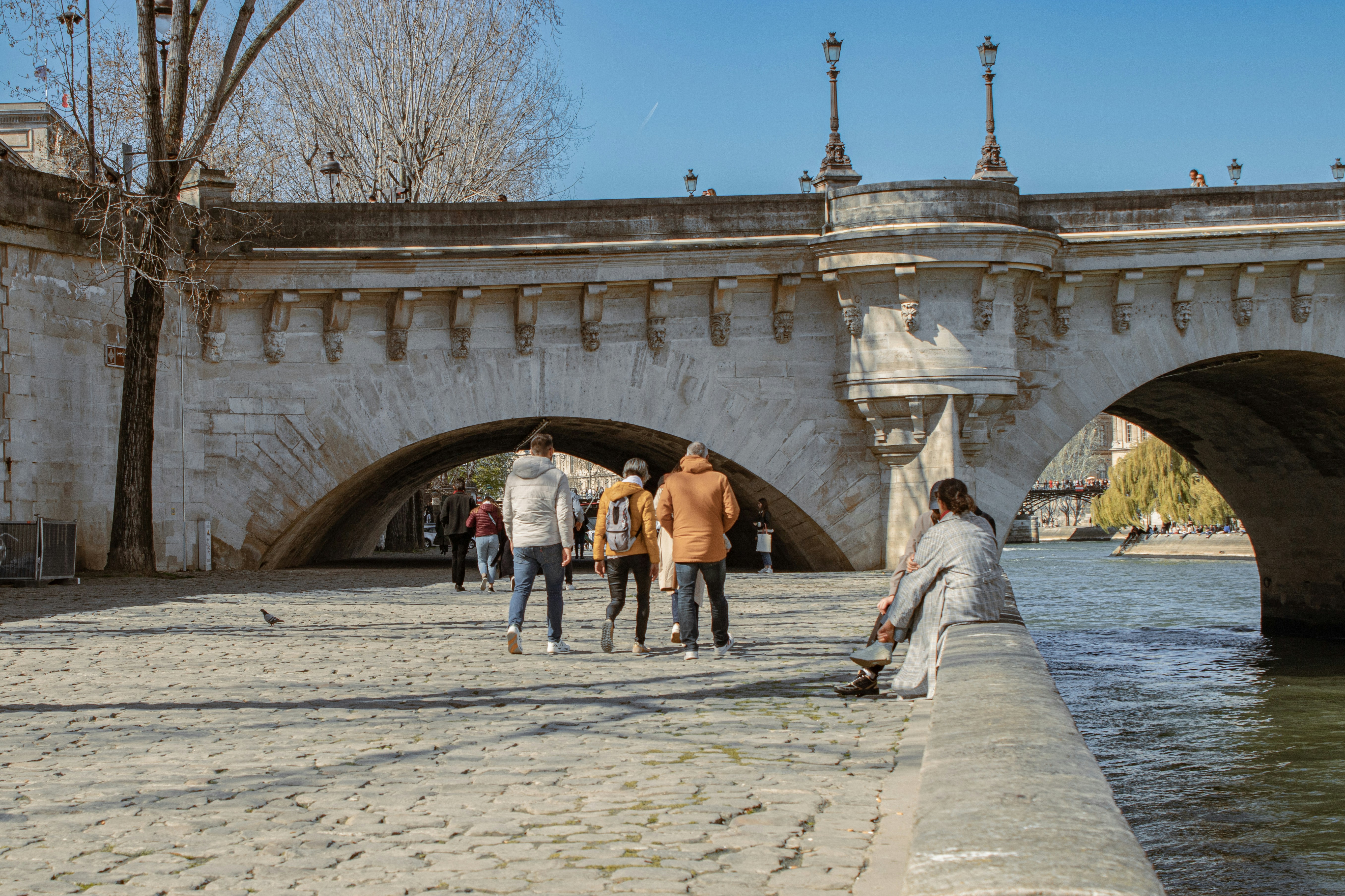 People walking along a cobblestone path beside a river, with a stone bridge in the background under a clear blue sky.