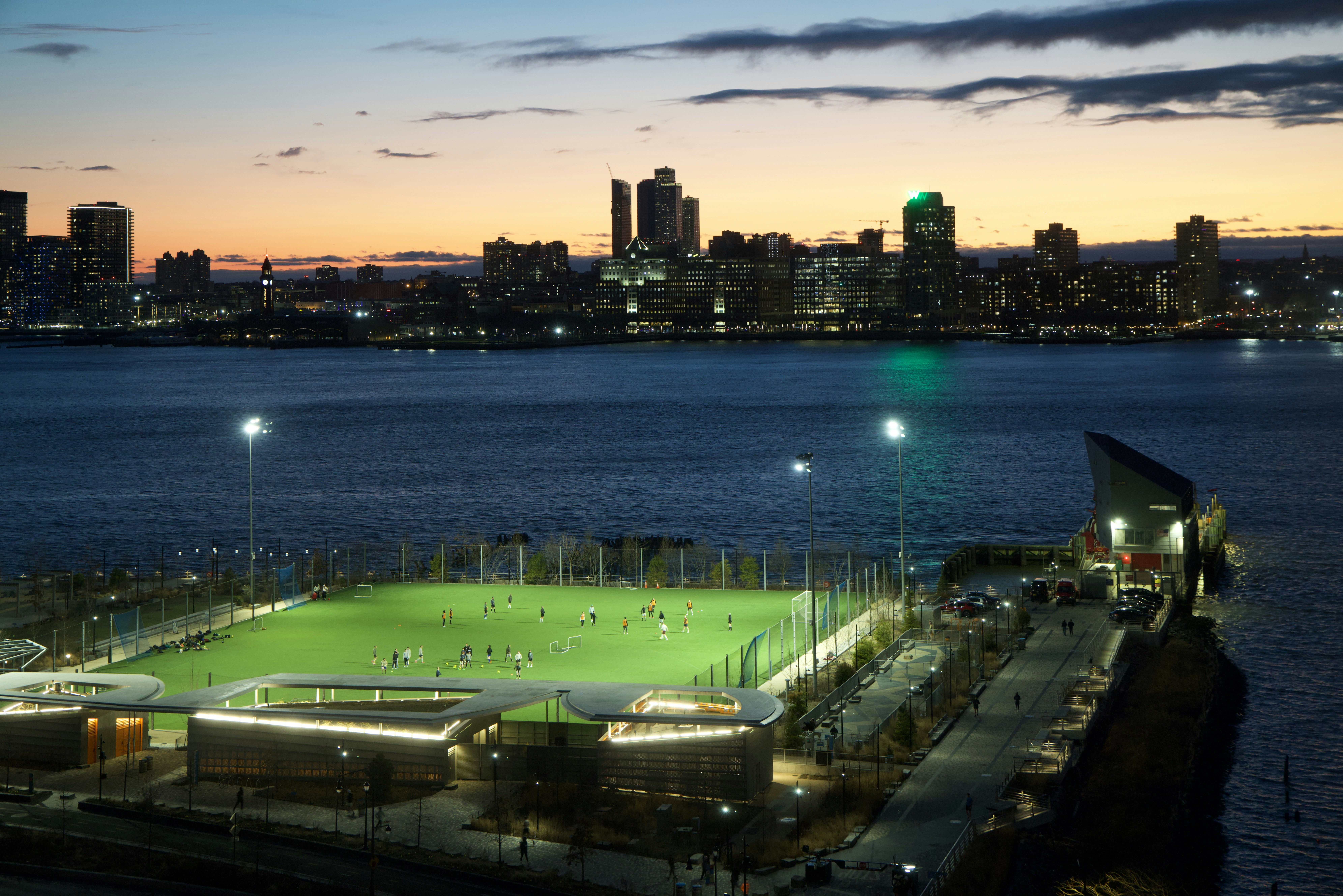 Soccer field illuminated at dusk beside the Hudson River with New Jersey skyline in the distance.