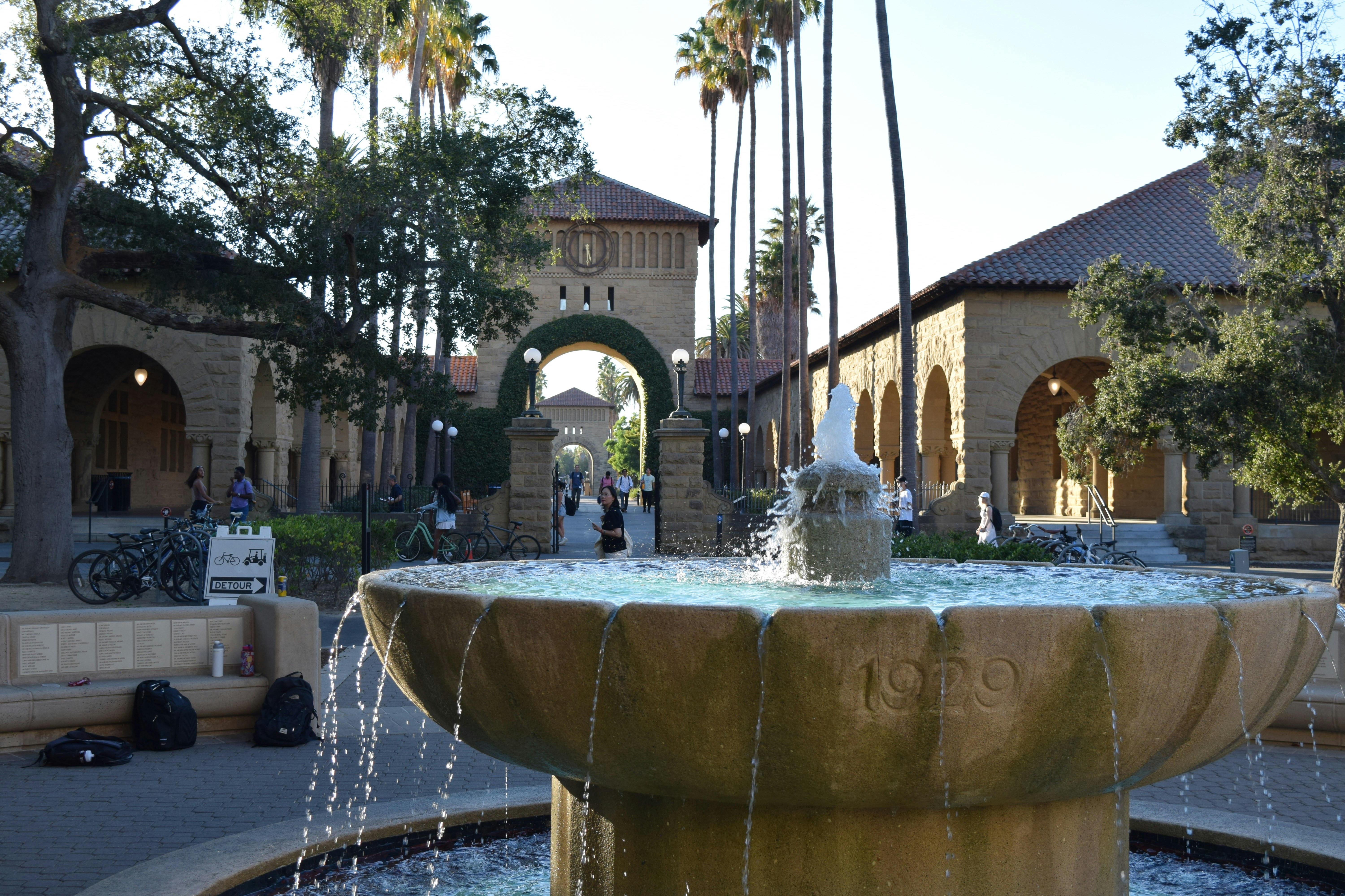 Stone fountain with cascading water set against historic campus buildings and palm trees under clear sky.