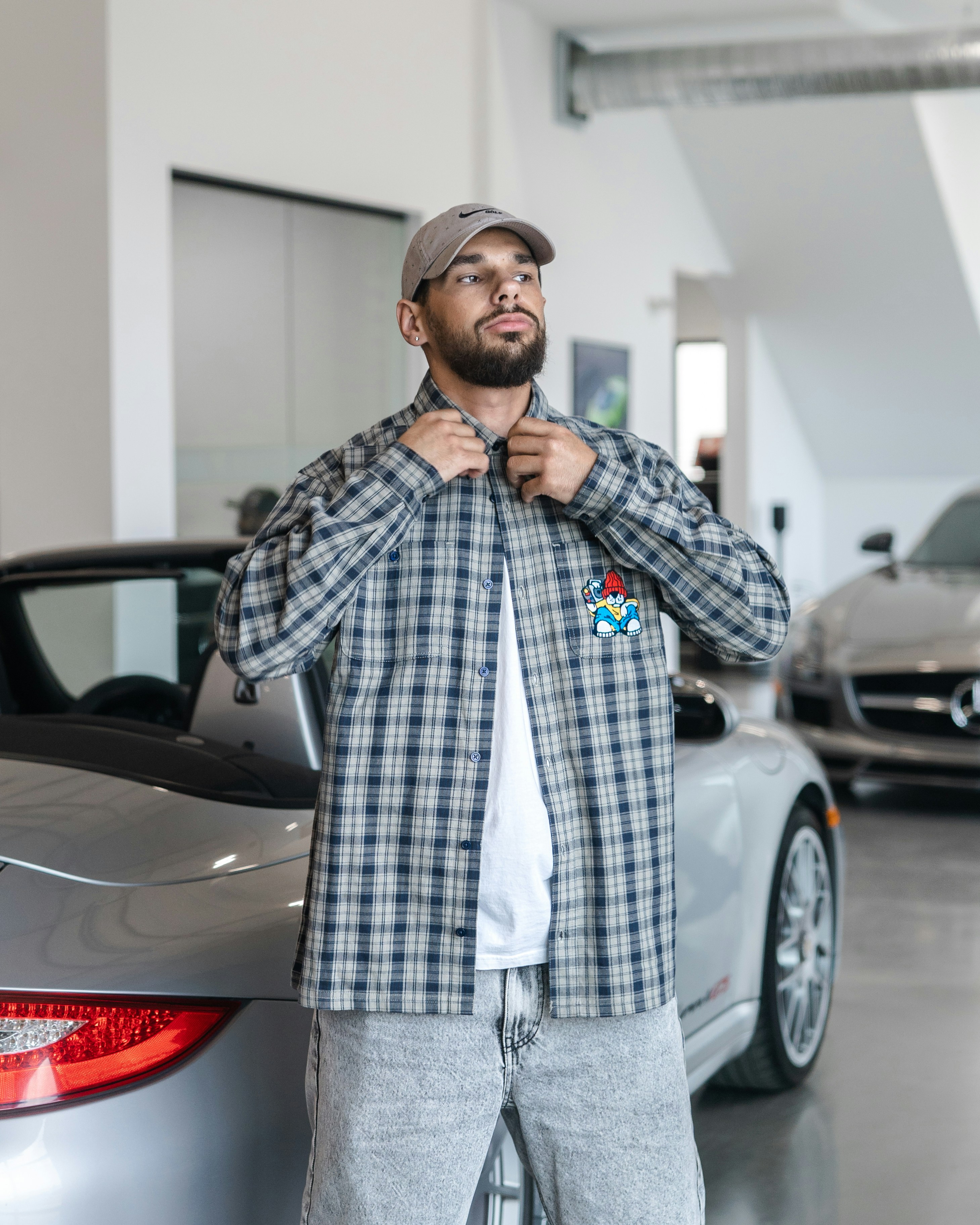 Man adjusting his shirt collar in a sleek garage, showcasing luxury vehicles in the background.
